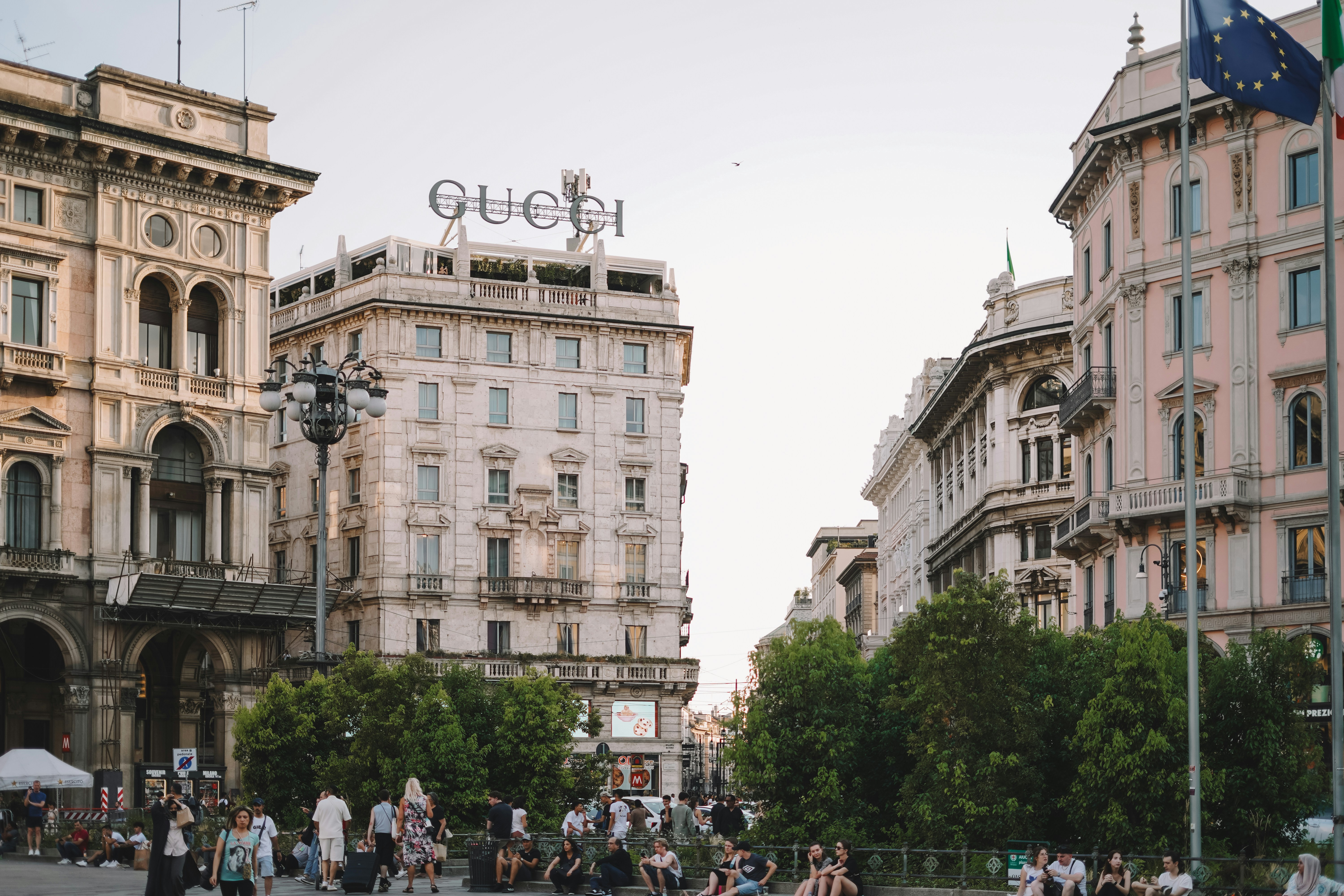 Historic buildings frame a bustling plaza in Milan, with a prominent Gucci sign highlighting the city's fashion culture. Crowds enjoy the vibrant atmosphere.