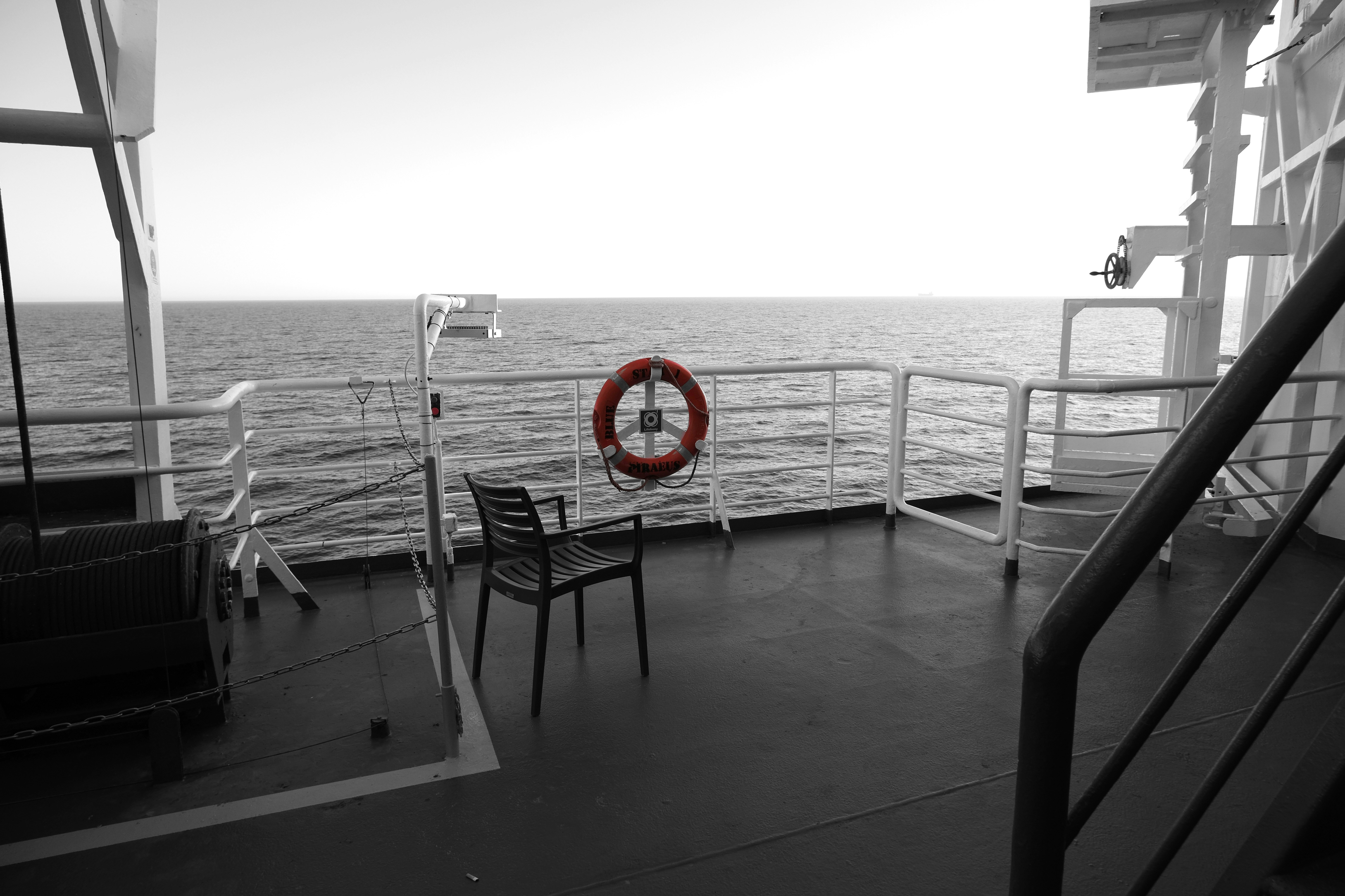 A lone chair beside a life buoy on a ship's deck overlooking the calm ocean. The scene conveys a sense of isolation and contemplation.