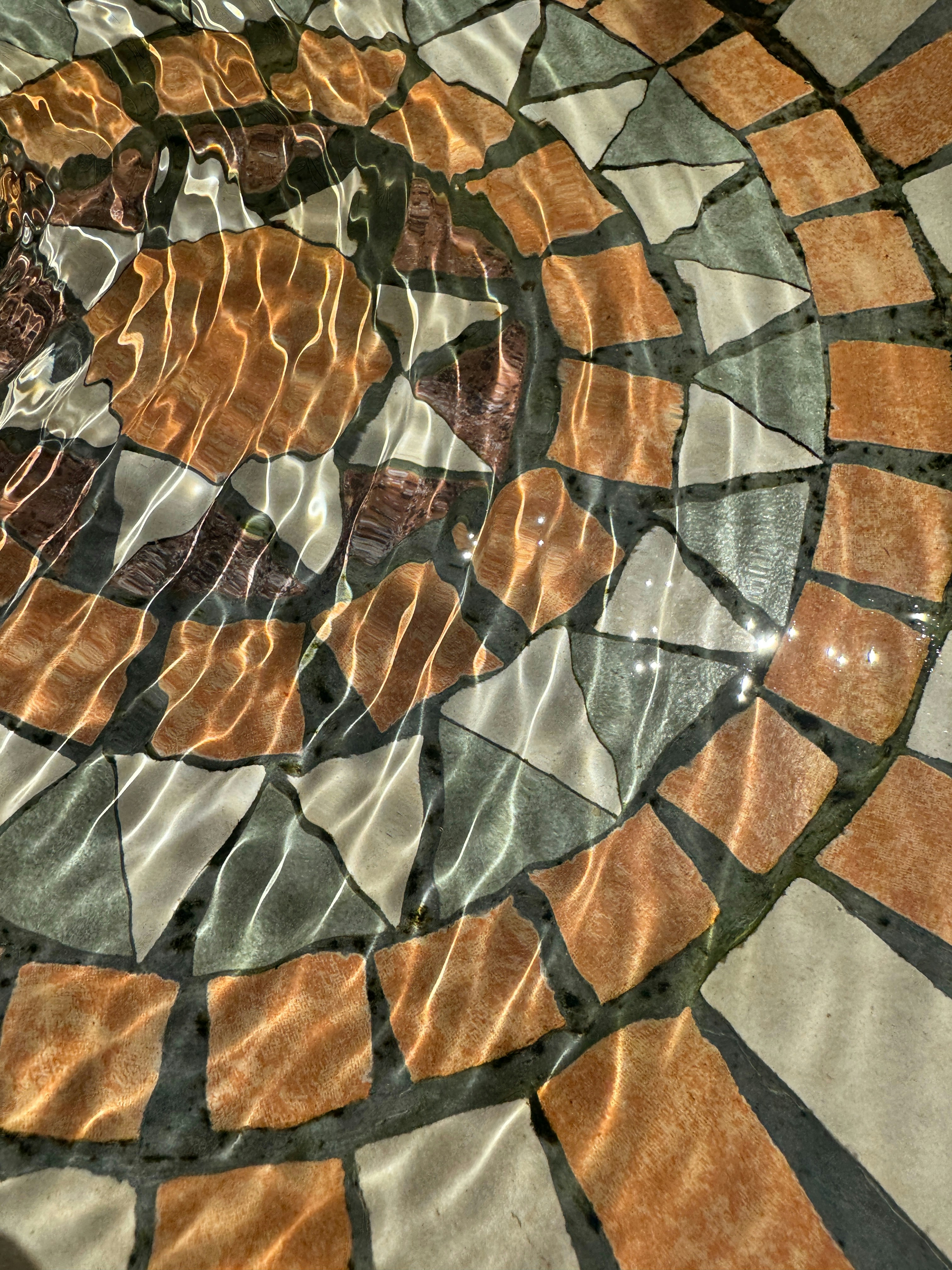 Close-up of a circular concentric tile mosaic in warm orange and cool gray stones with dark grout. Glossy, water-like reflections sweep over the surface.