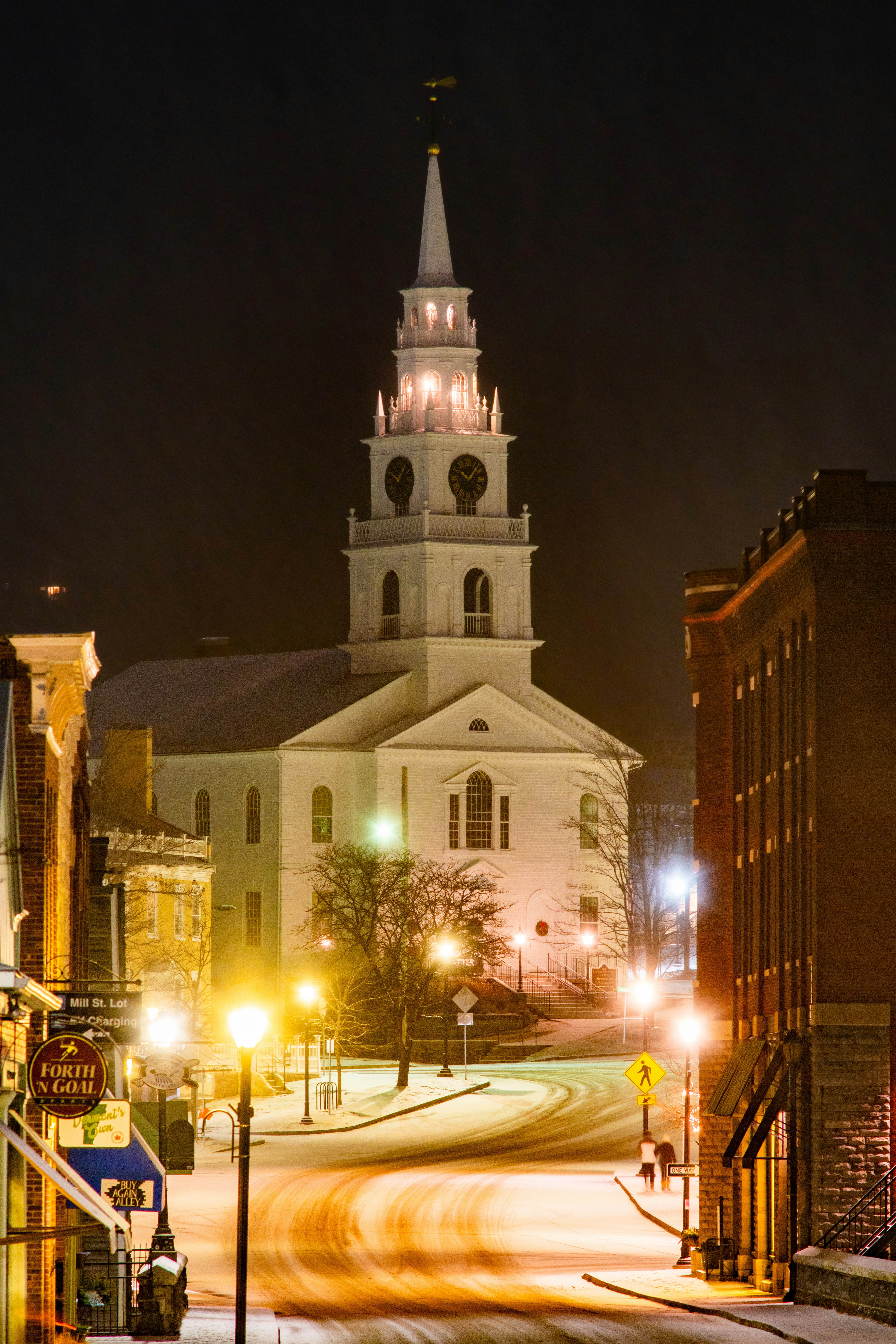 Historic church illuminated against a snowy street backdrop, with warm streetlights casting a glow. The scene captures the charm of a winter night in a quaint town.