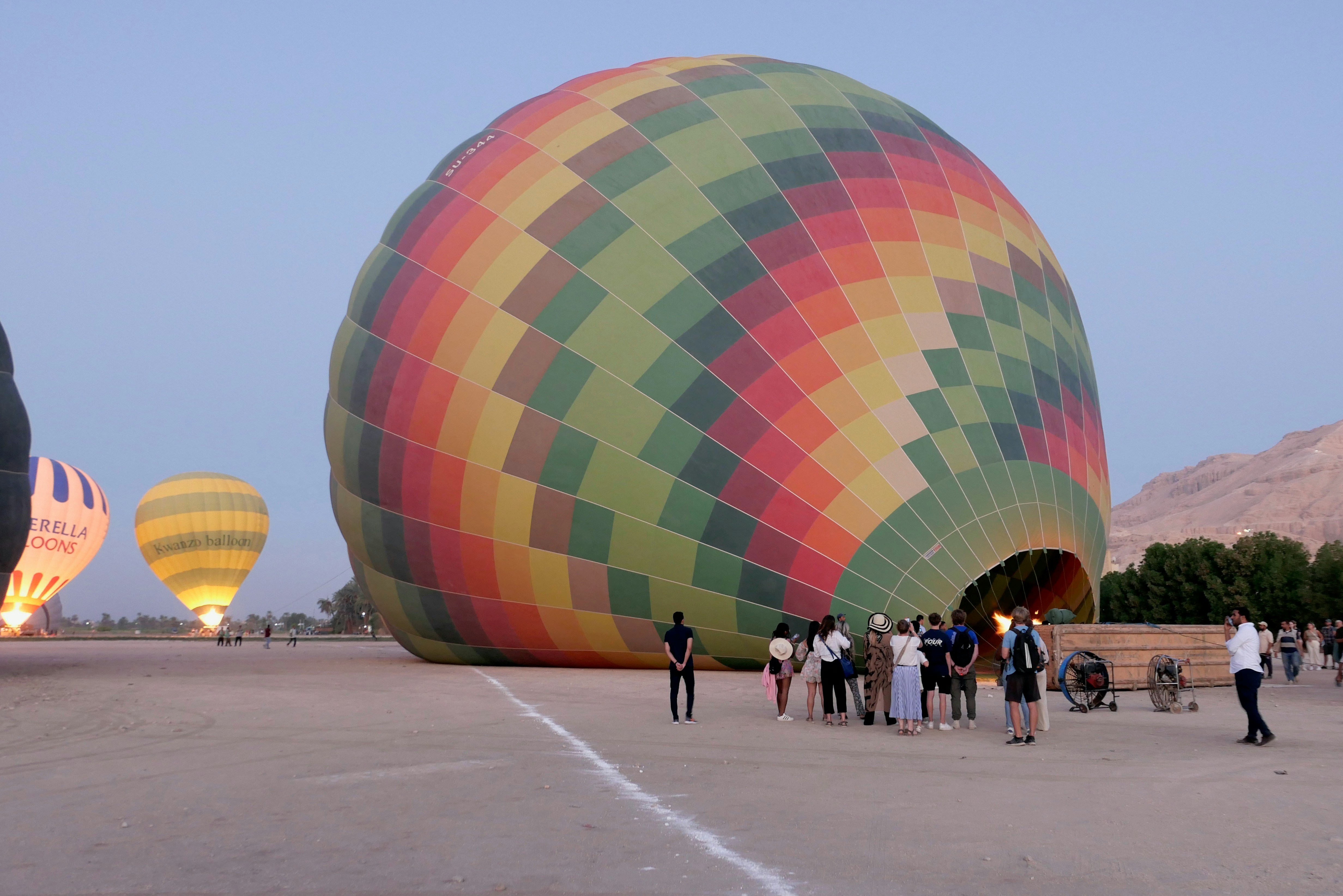 Photograph of a giant multicolored hot air balloon being inflated on a dusty field, with a group of observers nearby and other balloons in the background.
