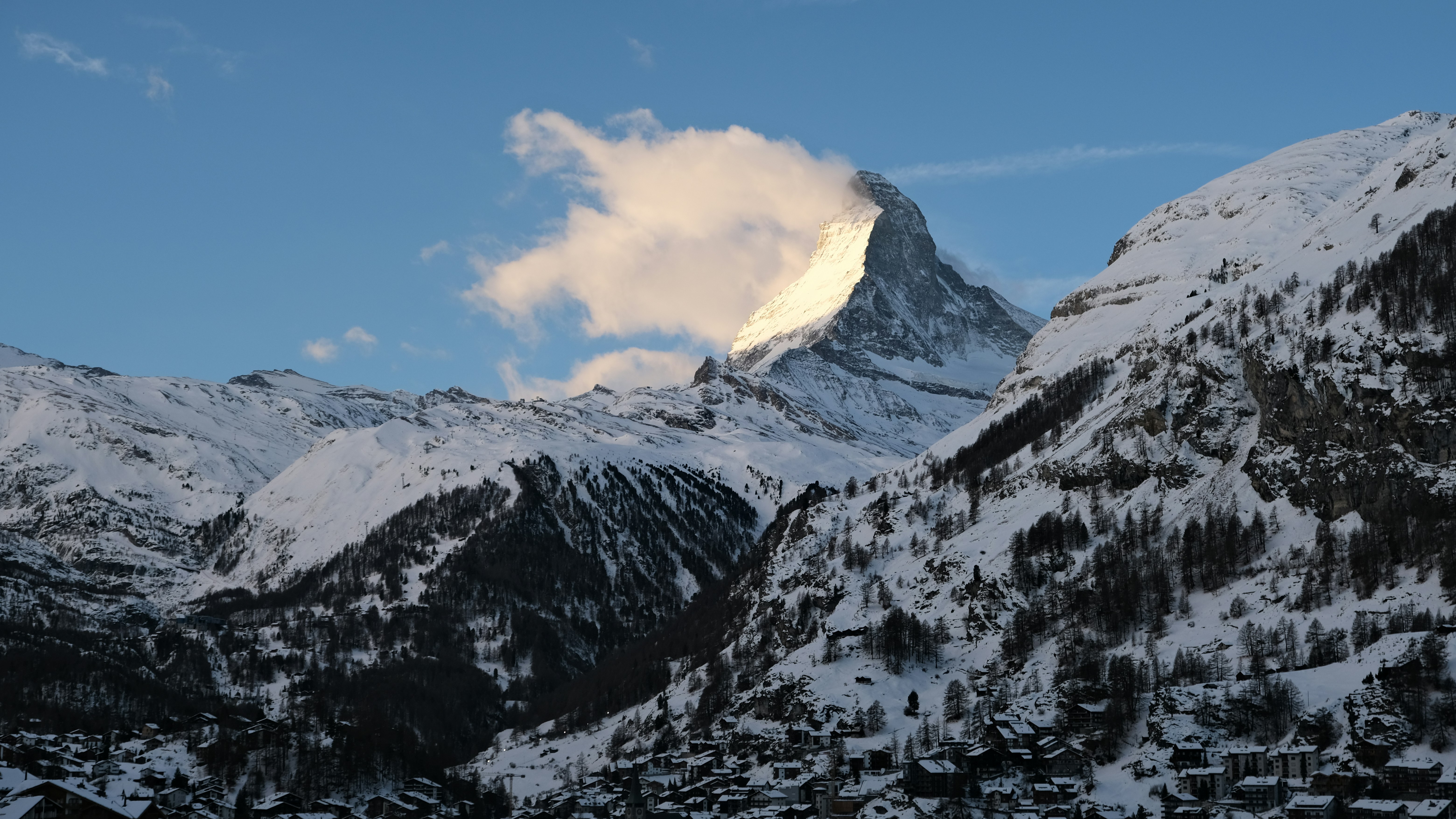 Snow-covered mountain peak kissed by sunlight, with a cloud hovering above against a clear blue sky.