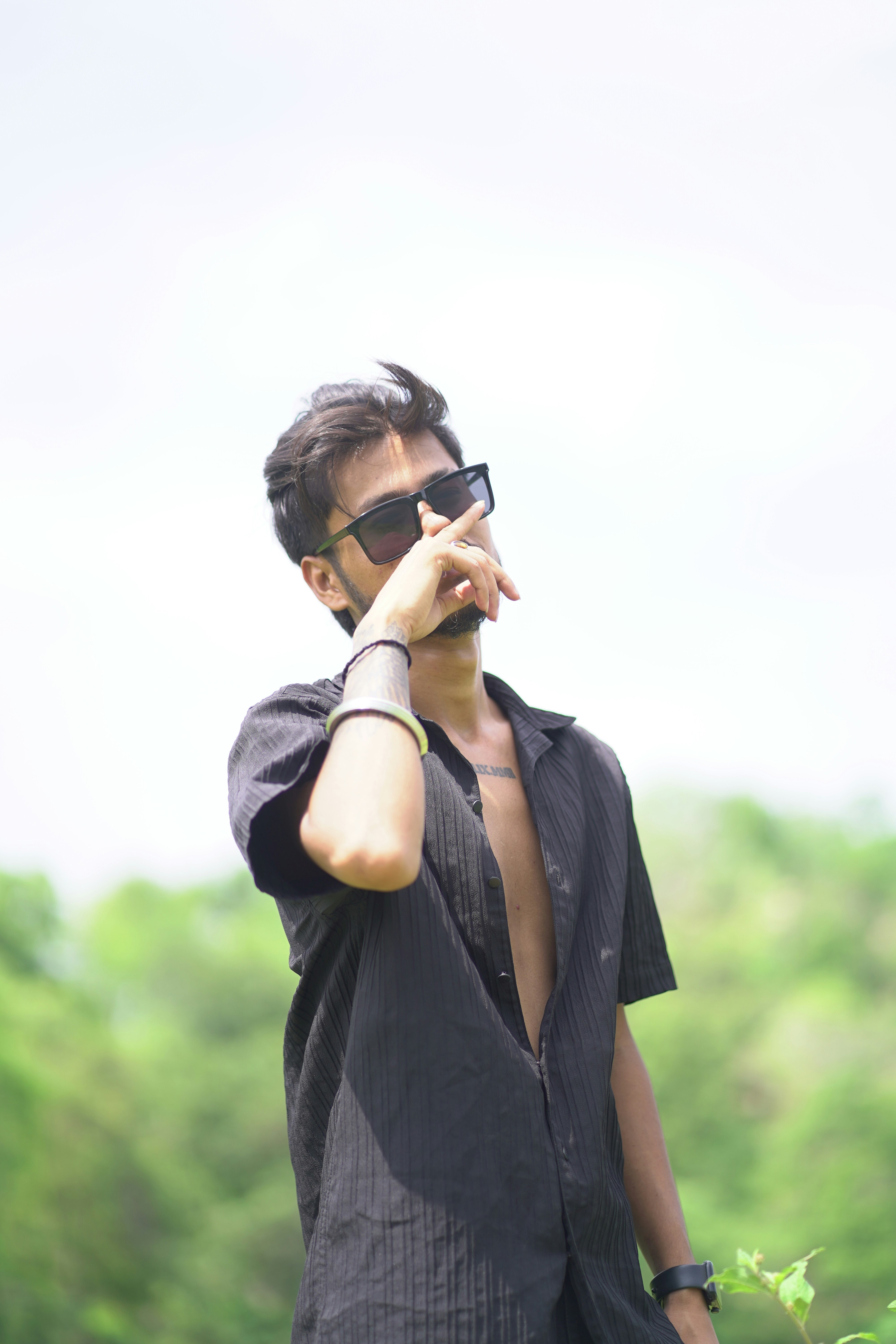 A young man in sunglasses poses thoughtfully against a lush green backdrop, embodying a moment of reflection.