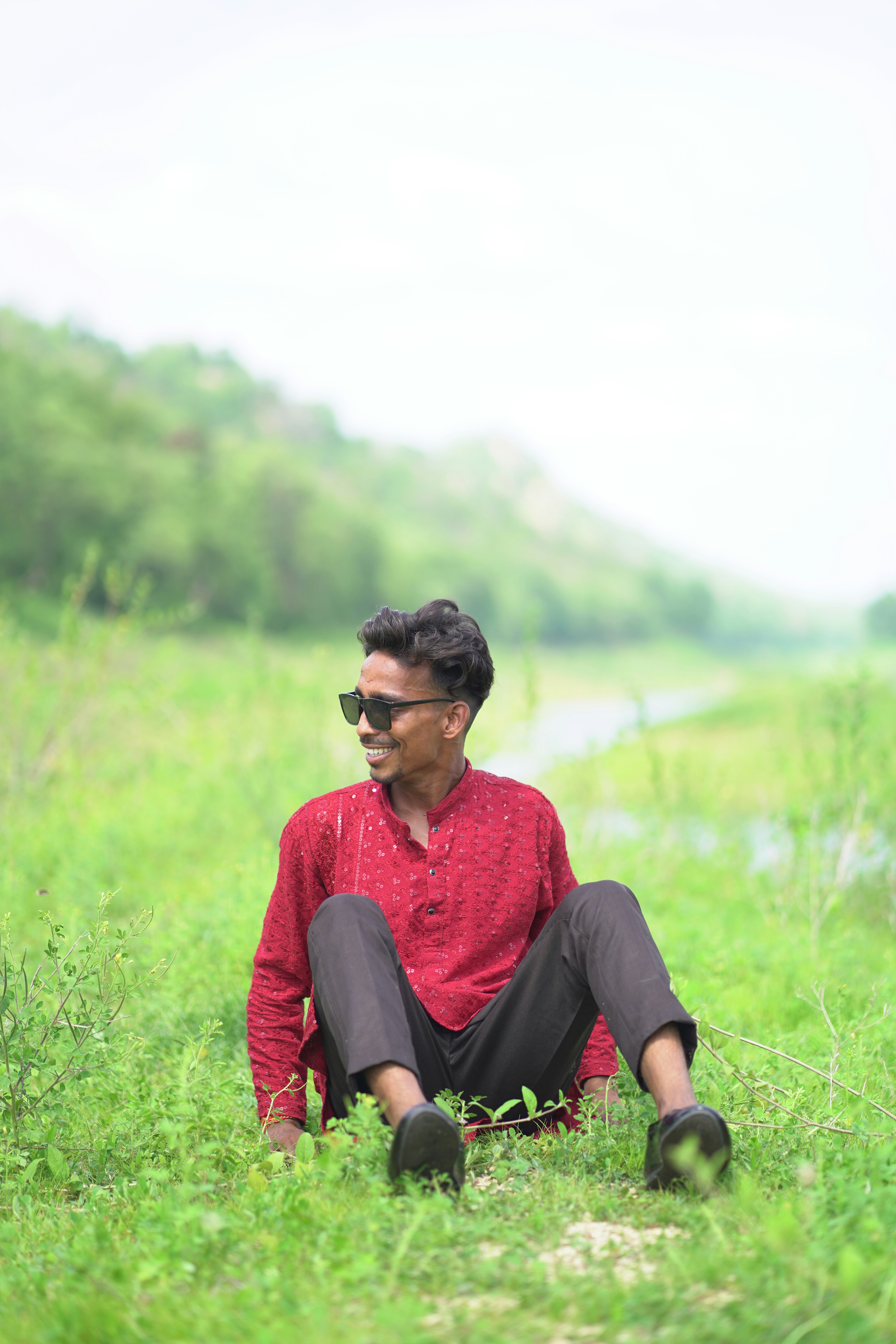 Young man in a red kurta sitting amidst lush greenery by a riverbank, radiating joy and calmness.