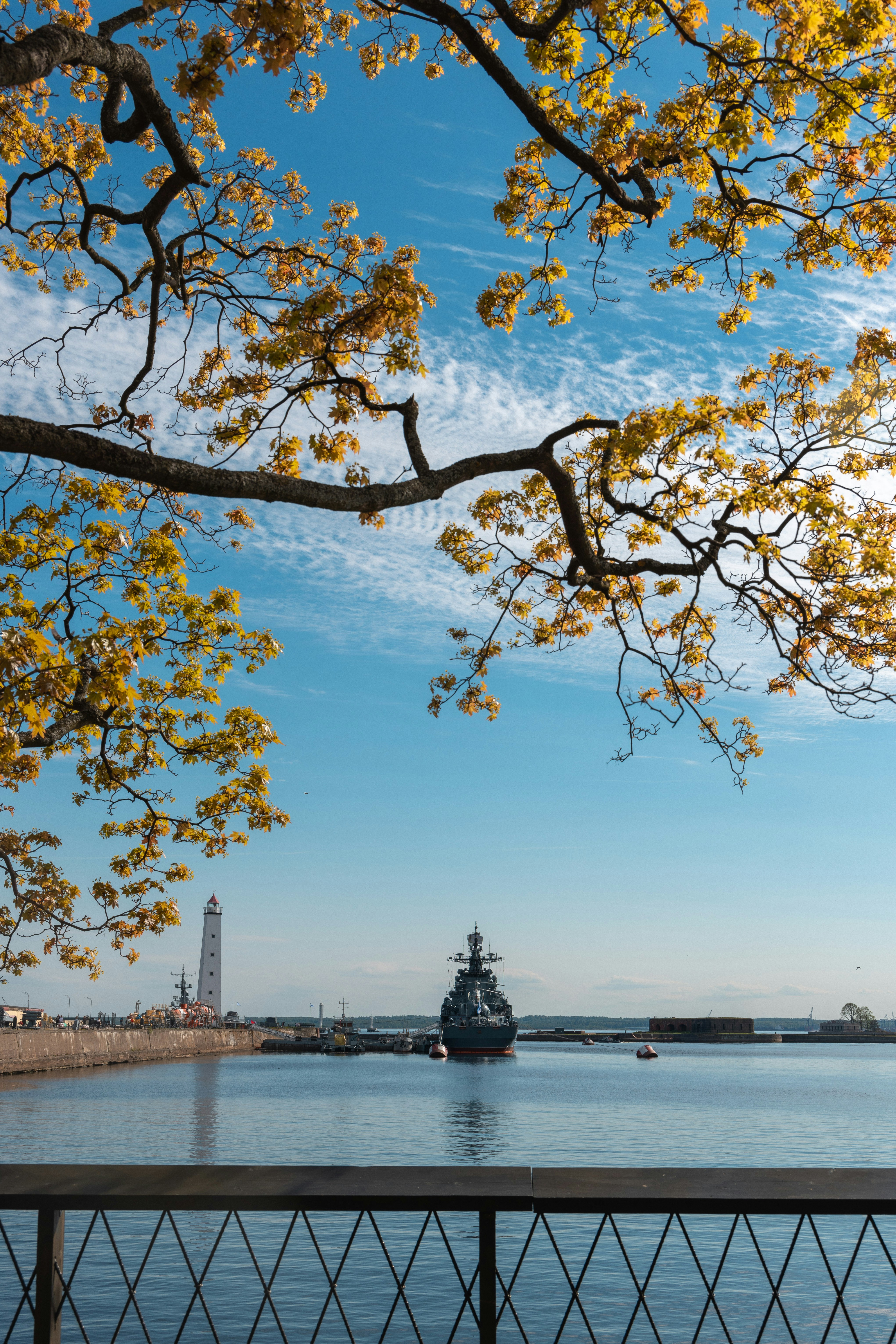 A naval ship anchored in a tranquil harbor, framed by vibrant autumn leaves and a distant lighthouse. The clear blue sky enhances the serene atmosphere.