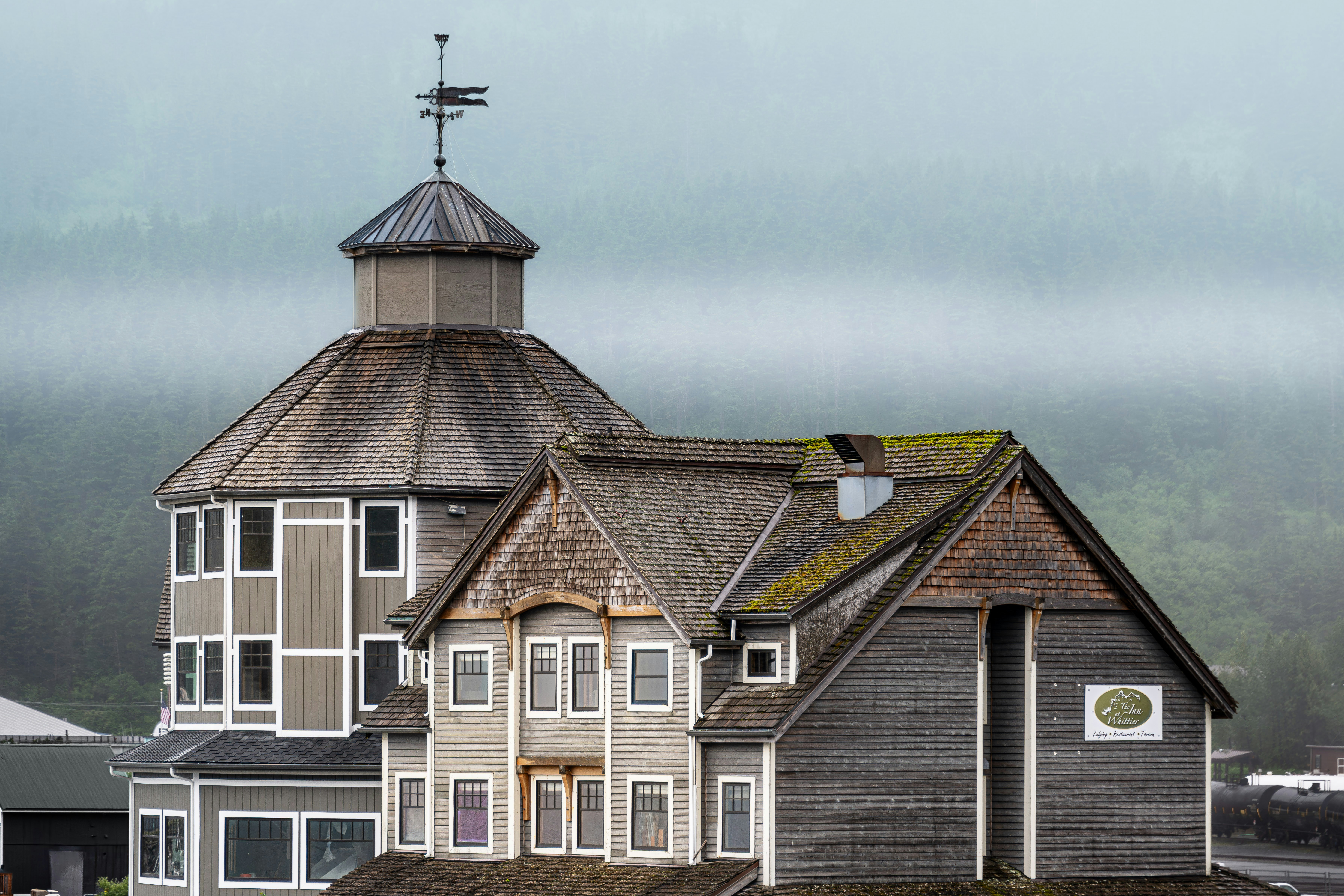 Wooden inn with weather vane, set against misty forest backdrop in Alaska.