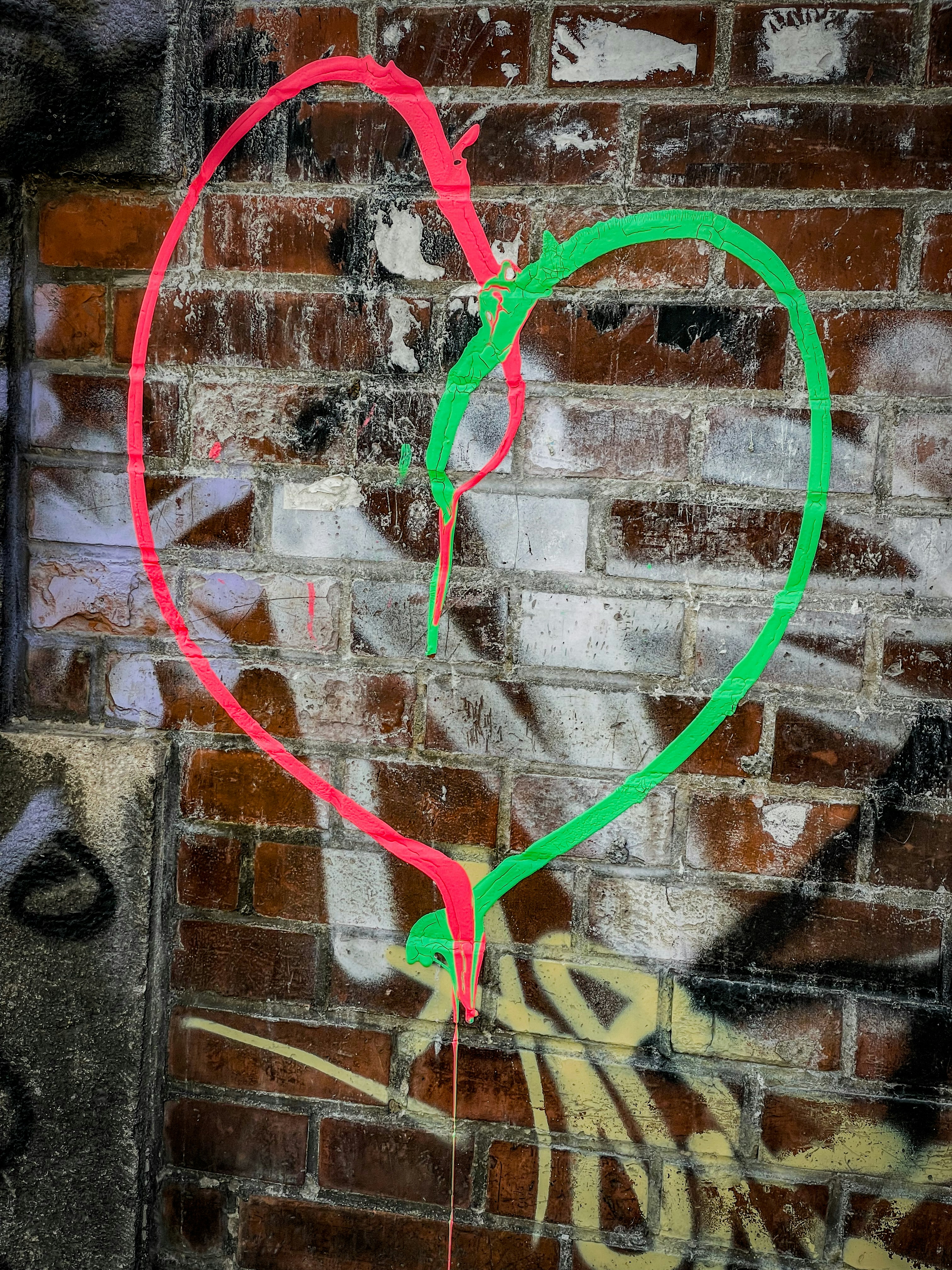 The image depicts a piece of street art featuring a heart shape painted on a brick wall. The heart is split into two halves, one in bright red and the other in vibrant green, with paint dripping down the wall. The background consists of a mix of bricks and other graffiti, creating a textured and urban aesthetic. The colorful heart stands out against the gritty backdrop, showcasing a blend of creativity and urban decay.