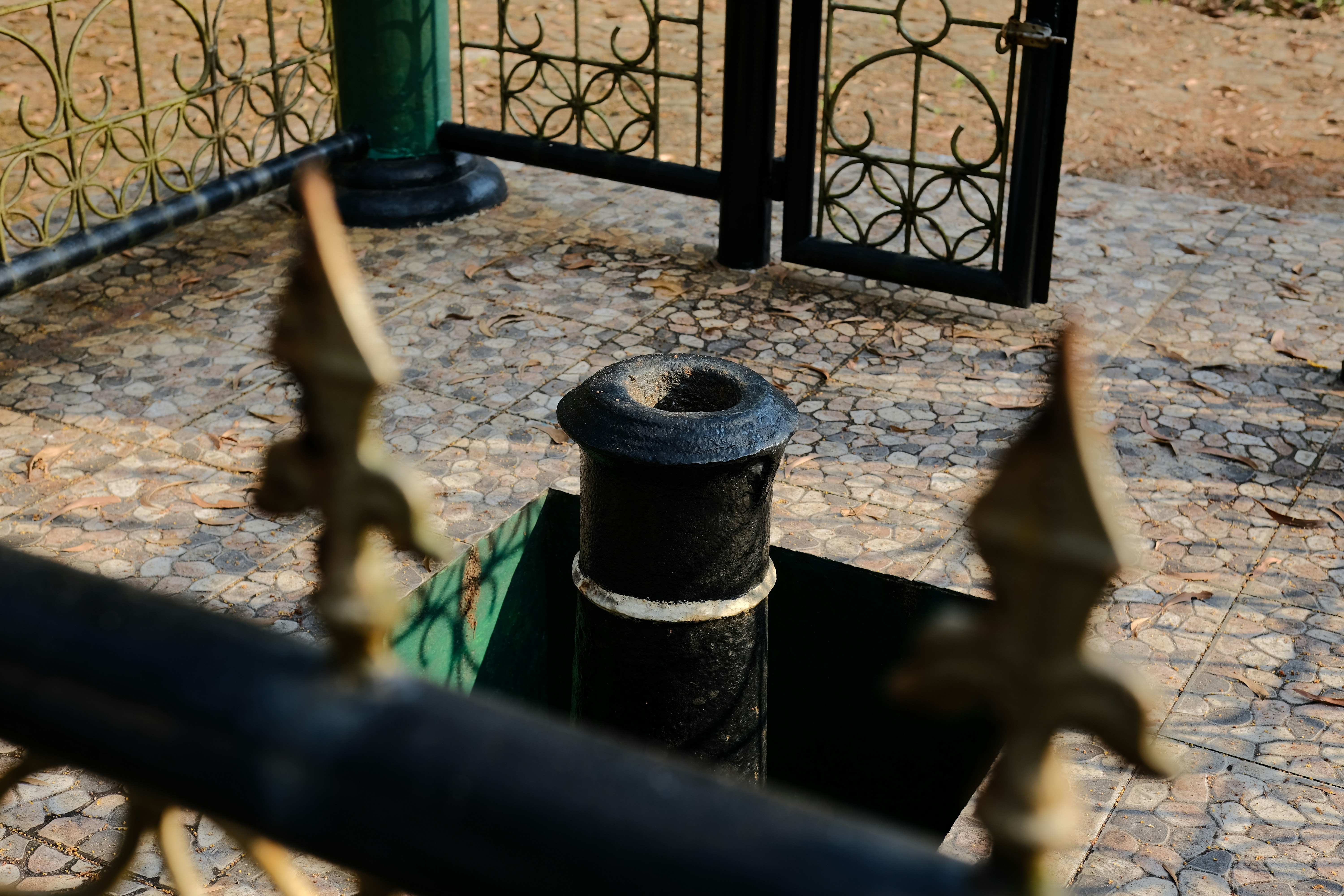 This photo shows a black cannon standing upright on a mosaic-tiled floor, surrounded by a decorative iron fence. The cannon has two white rings around its body and looks well-maintained, with some dry leaves scattered around and sunlight illuminating the area.