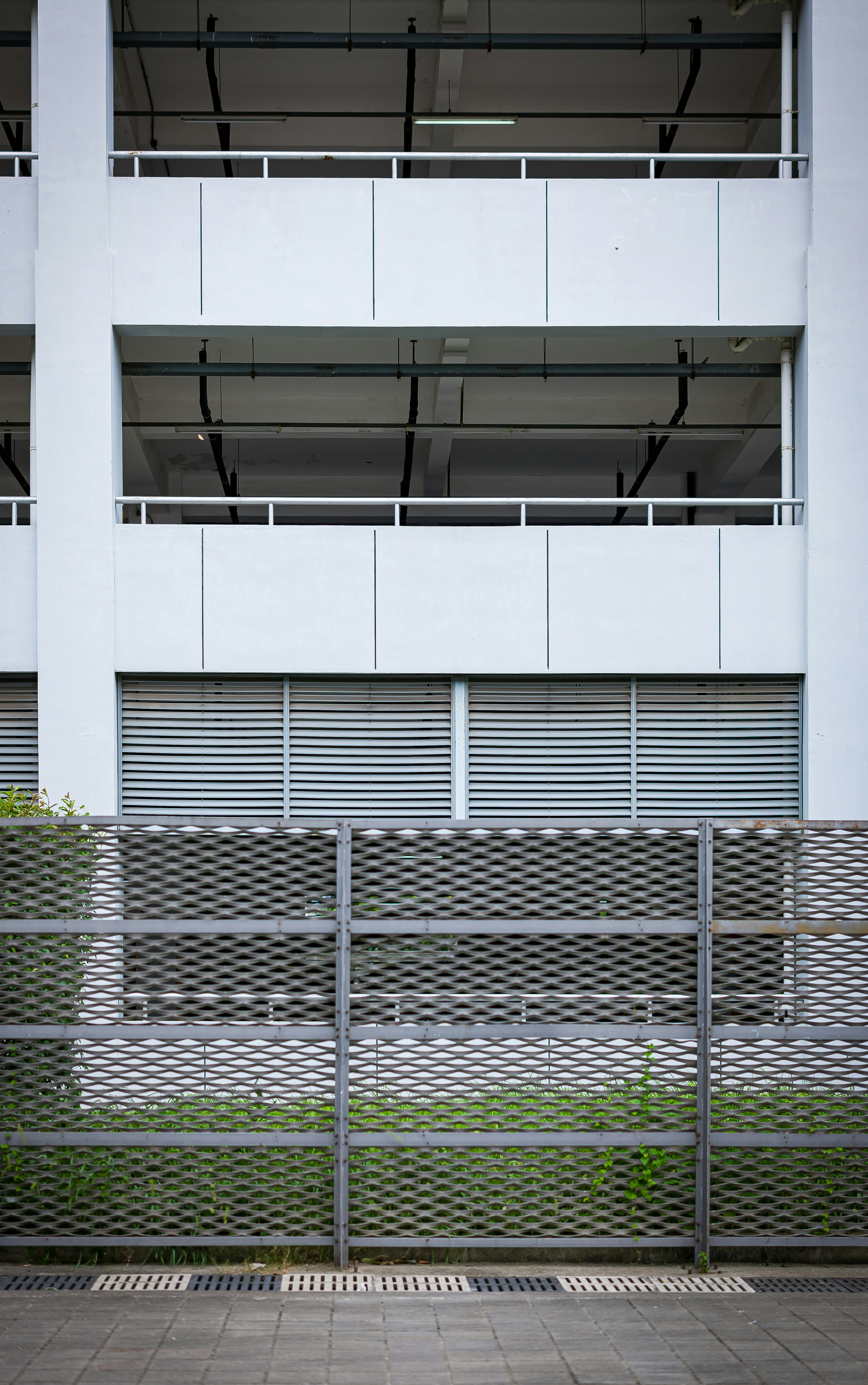 Architectural facade of a multi-level parking structure with white panels and horizontal louvers, seen behind a perforated metal fence. Clean lines and muted tones emphasize industrial geometry.