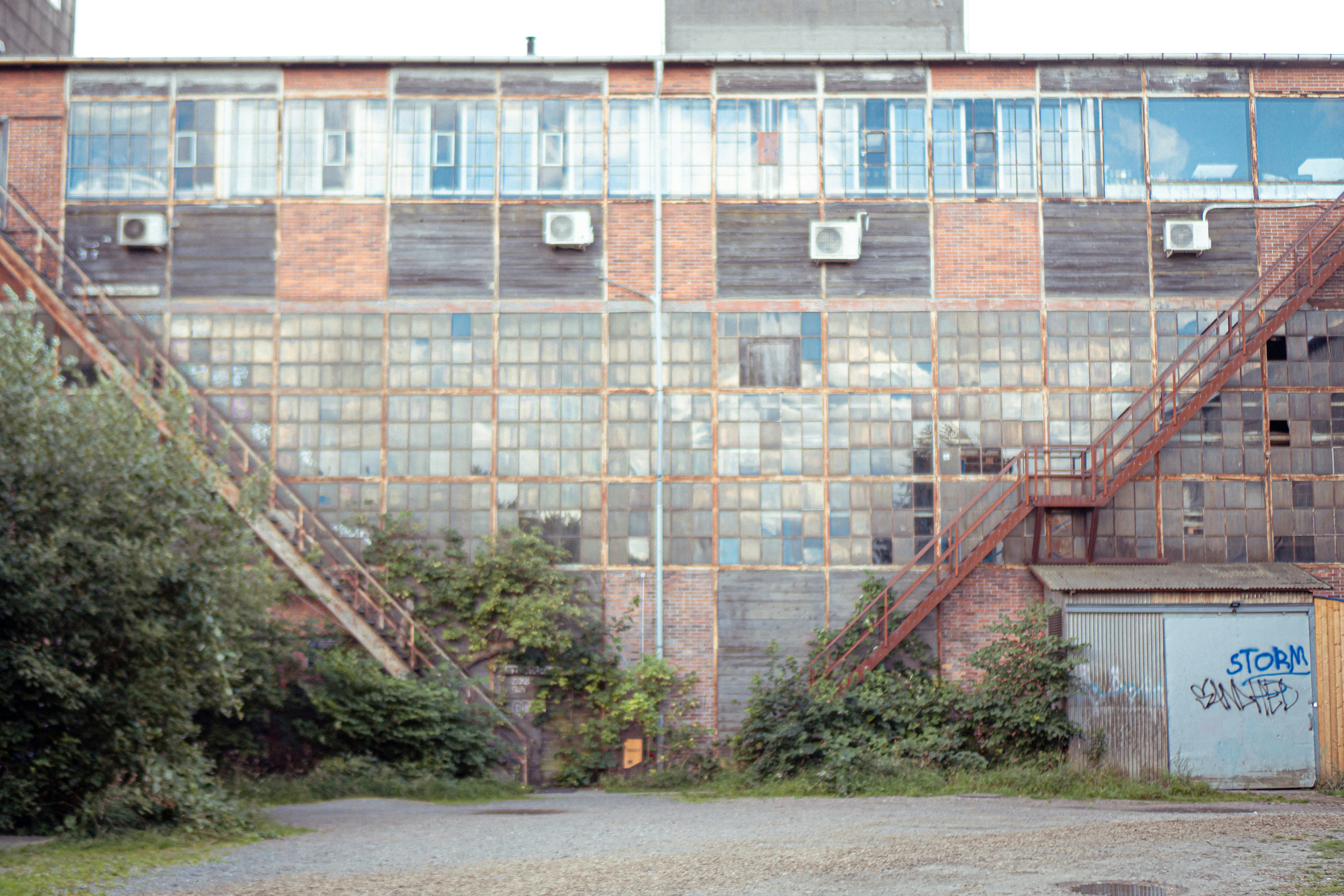Abandoned industrial building featuring a patchwork of windows and rusting staircases, surrounded by overgrown vegetation.
