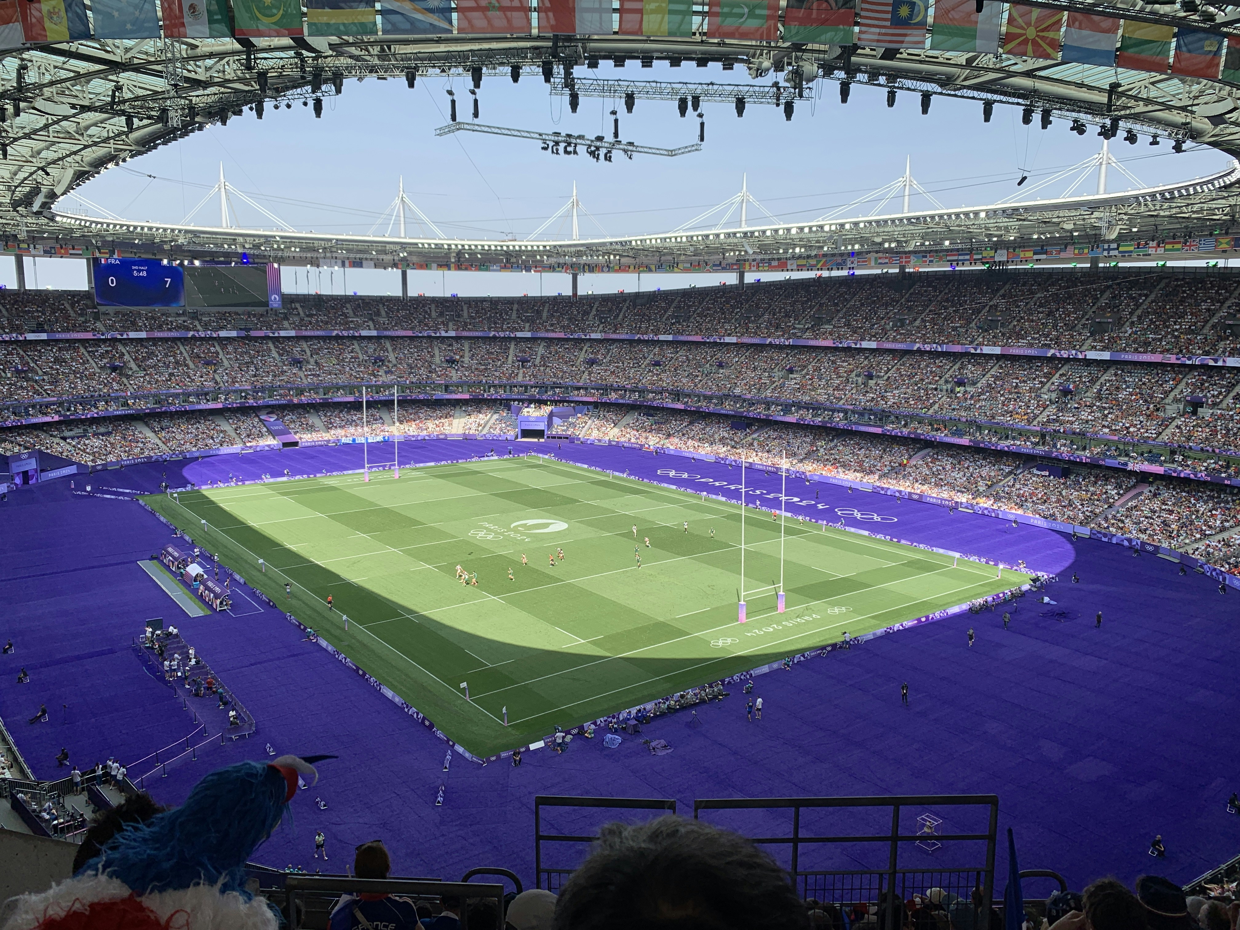 Aerial view of a rugby match in a packed stadium, showcasing vibrant purple turf and enthusiastic spectators under a clear sky.