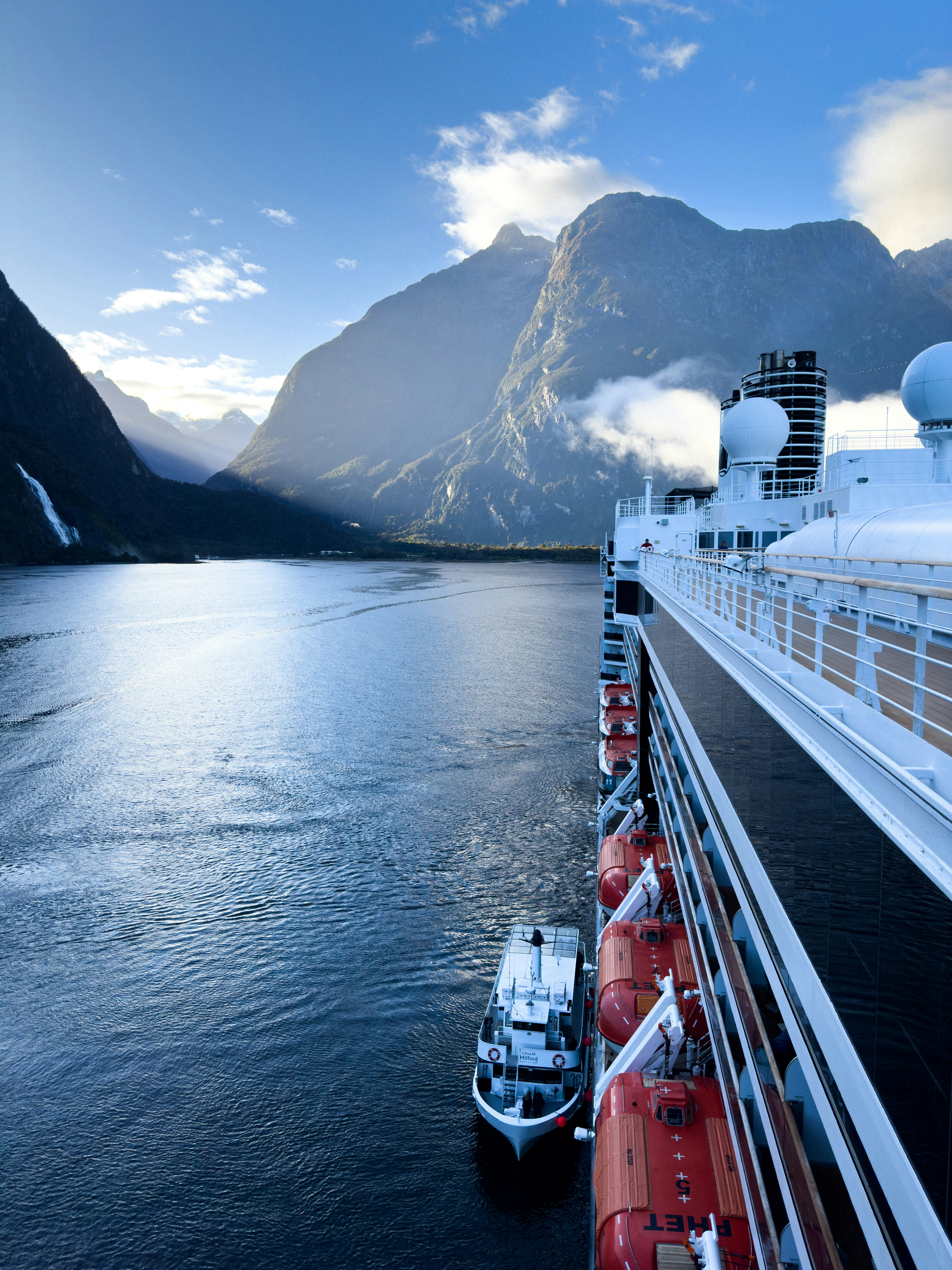 A cruise ship glides through tranquil waters, framed by towering mountains and soft clouds. The scene captures the harmony of nature and maritime adventure.