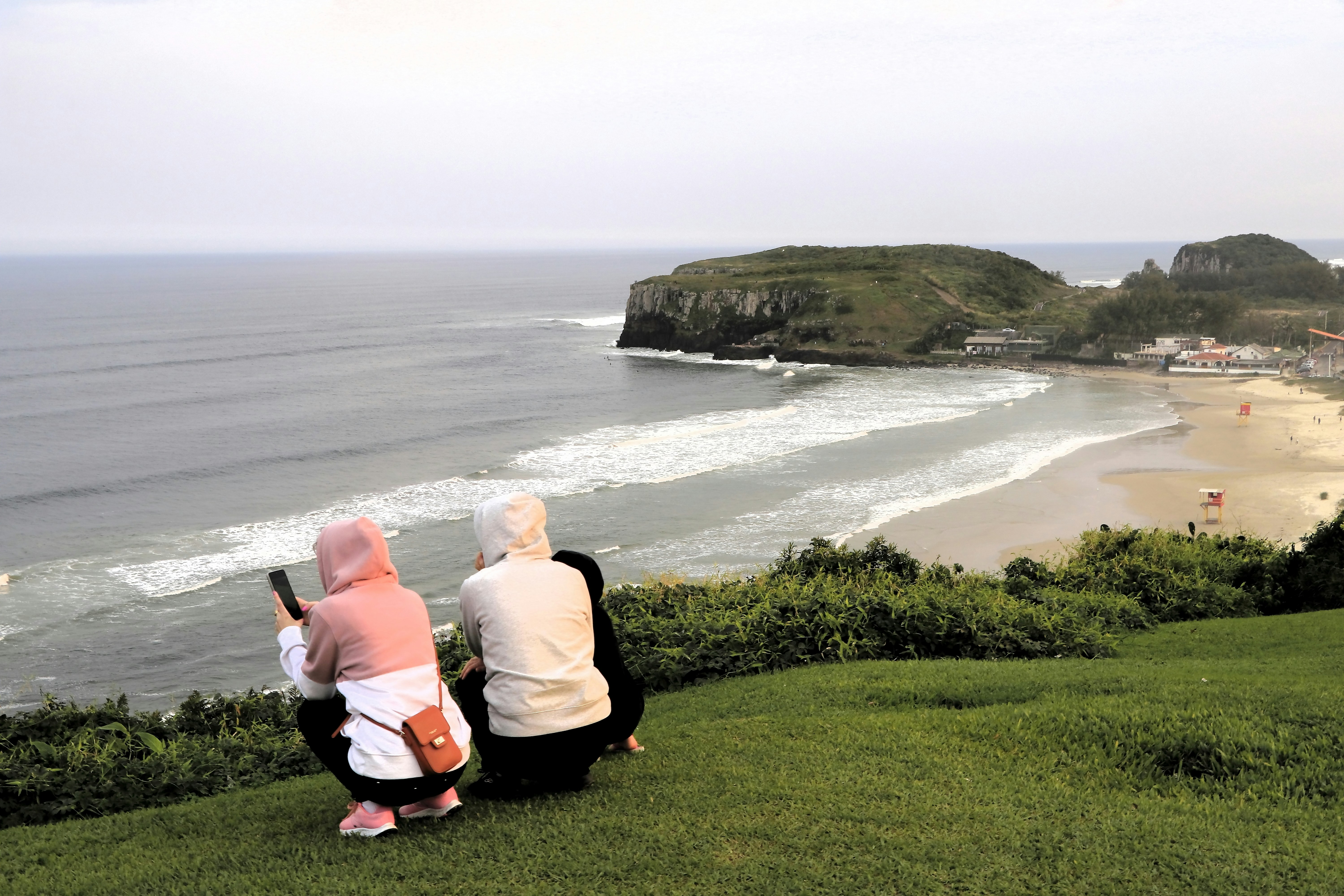 Two people sit on grass overlooking a coastal landscape with waves crashing against distant cliffs under a cloudy sky.