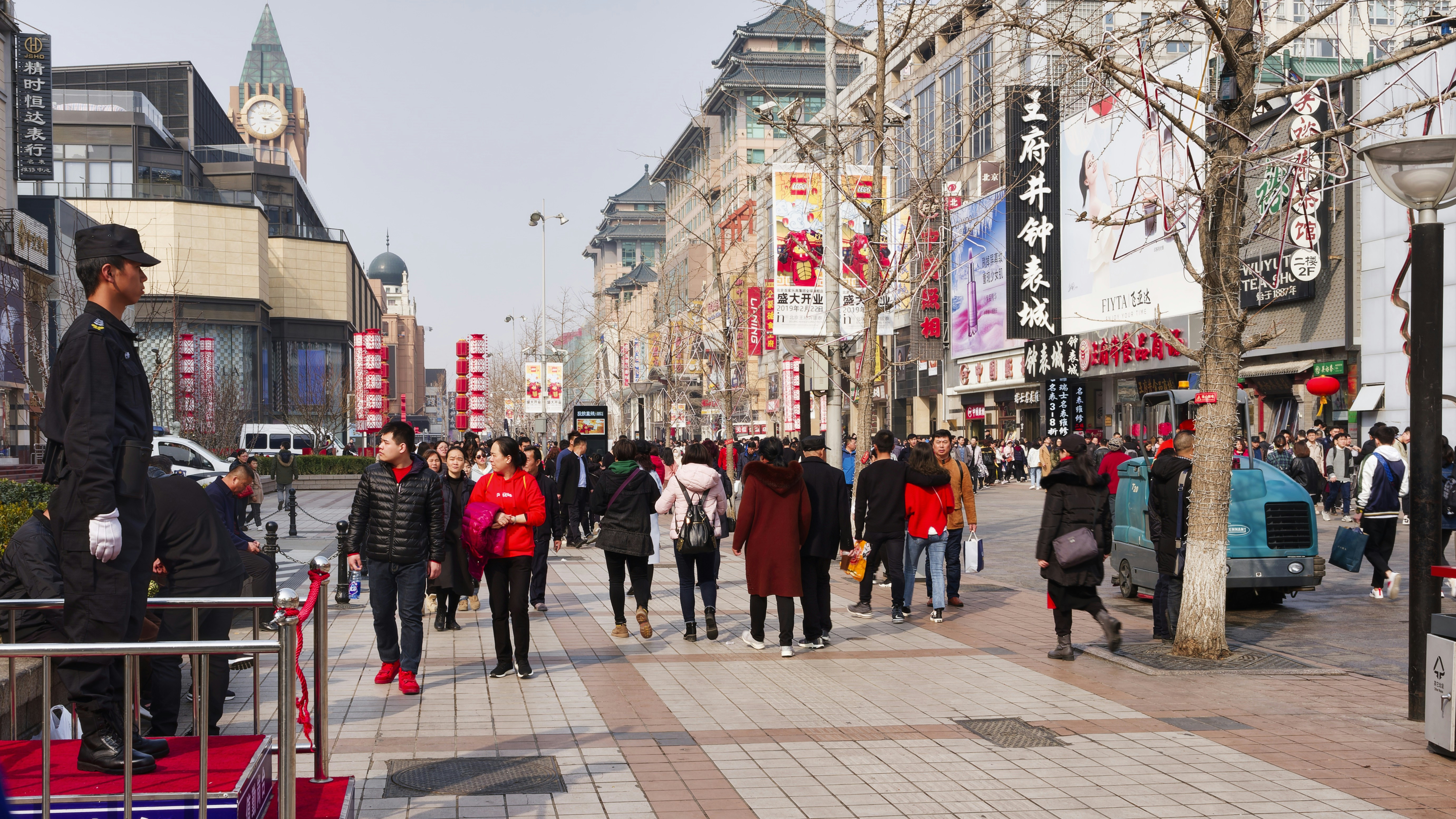 Pedestrians stroll along a lively city street lined with colorful storefronts and advertisements, while a security guard stands watchfully.