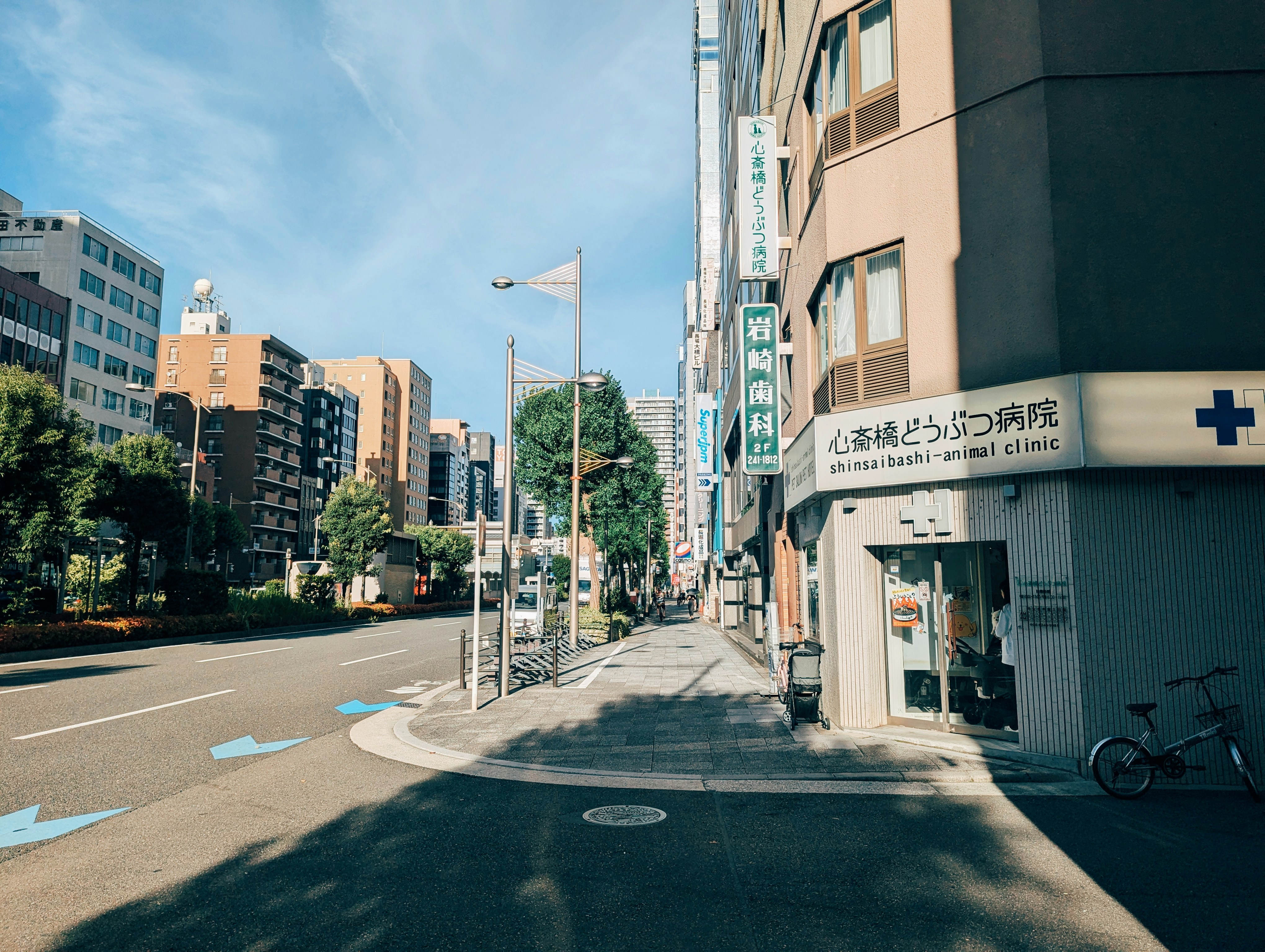 Sunlit city street with modern buildings and Japanese signage extends toward a distant vanishing point. A quiet sidewalk lines the right, with a storefront and parked bicycle.