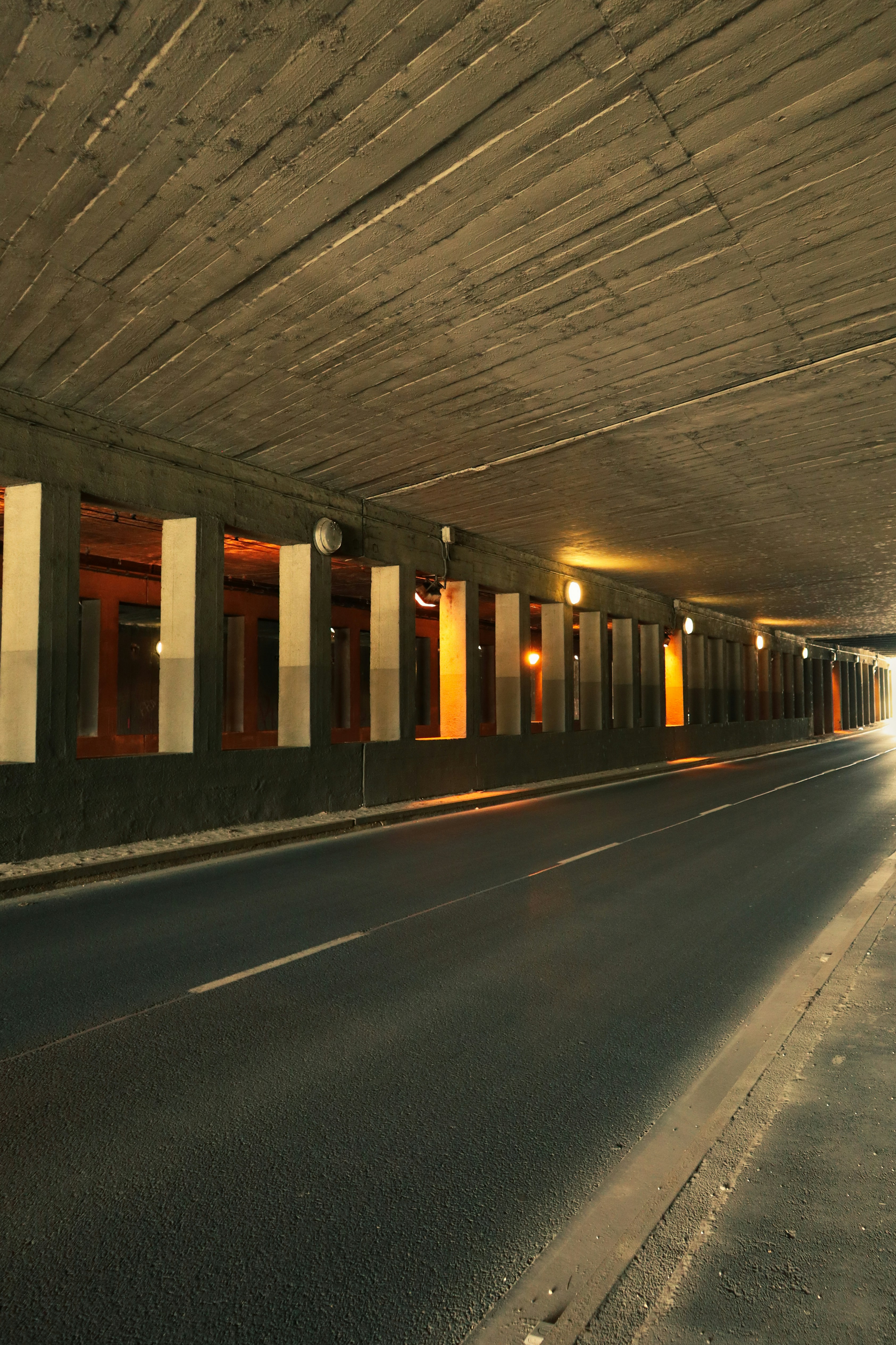 Dimly lit underpass with concrete pillars and warm lights, creating a moody atmosphere. The empty road leads into the distance.