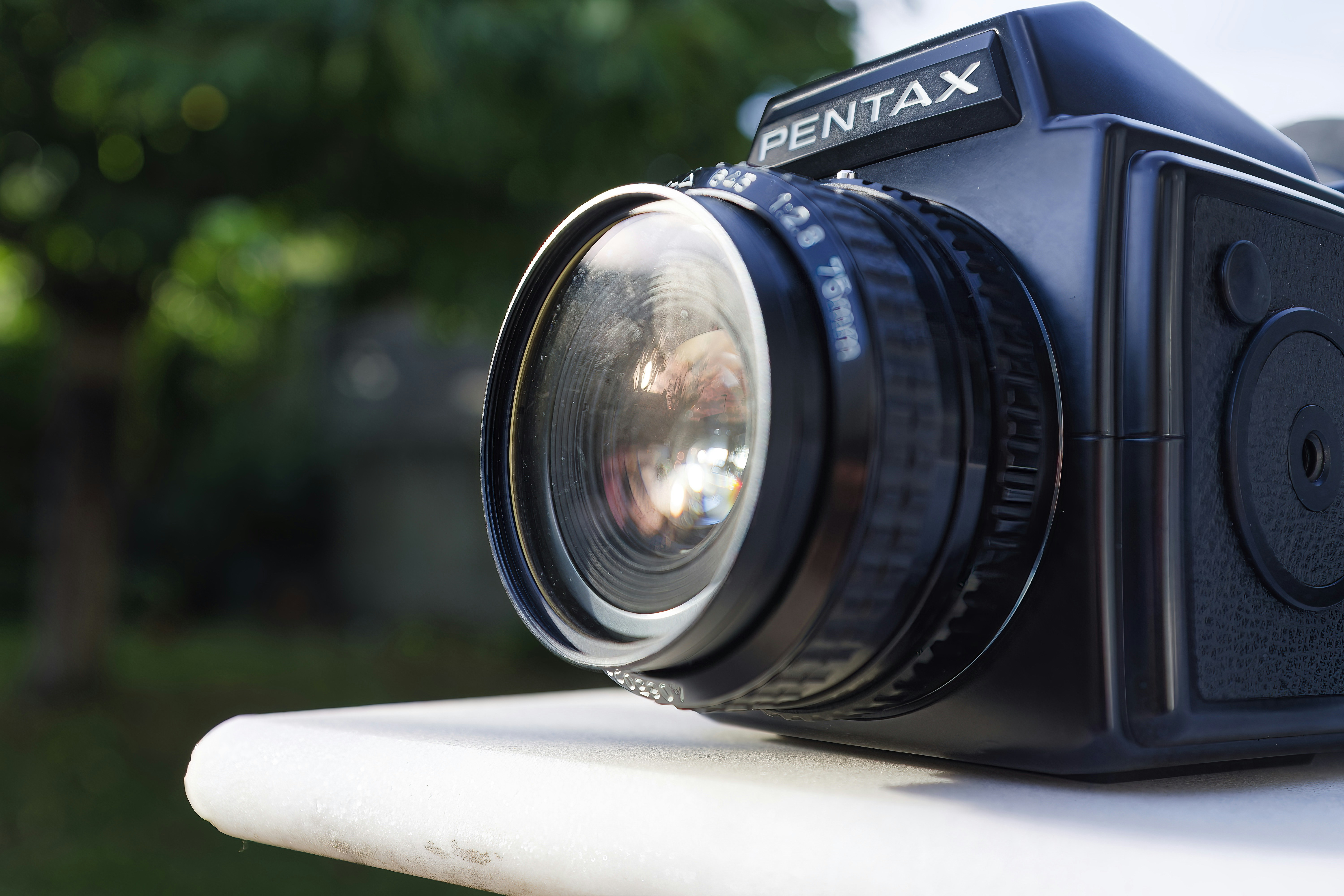Vintage Pentax camera on a white surface amidst lush green foliage and sunlight.