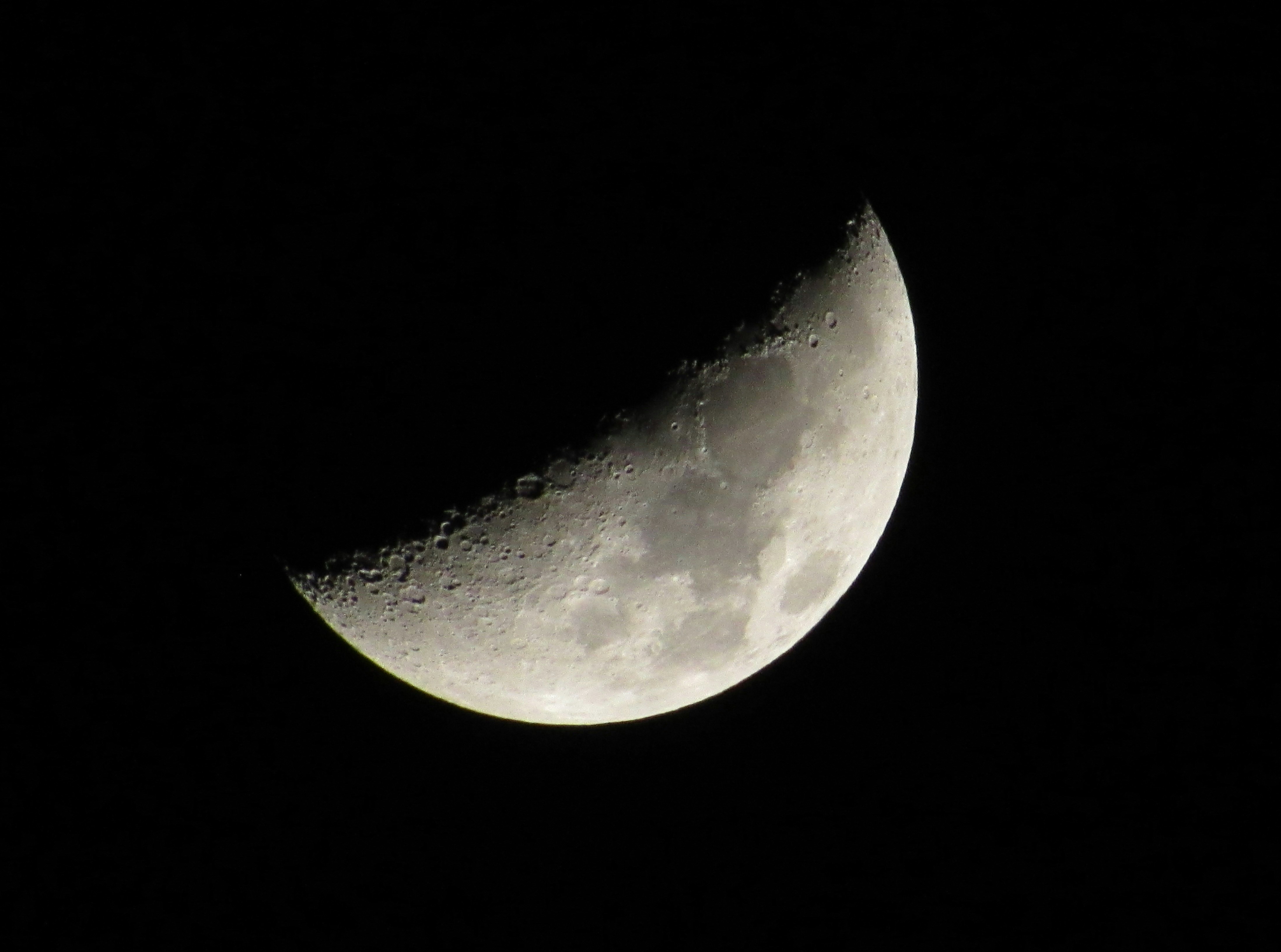 A close-up photo of a crescent moon in a dark night sky. The illuminated portion of the moon is a bright white, with visible craters and surface details. The dark side of the moon is faintly visible, with a soft, gray glow. The crescent shape is clear and defined, with a sharp terminator between the lit and unlit parts.