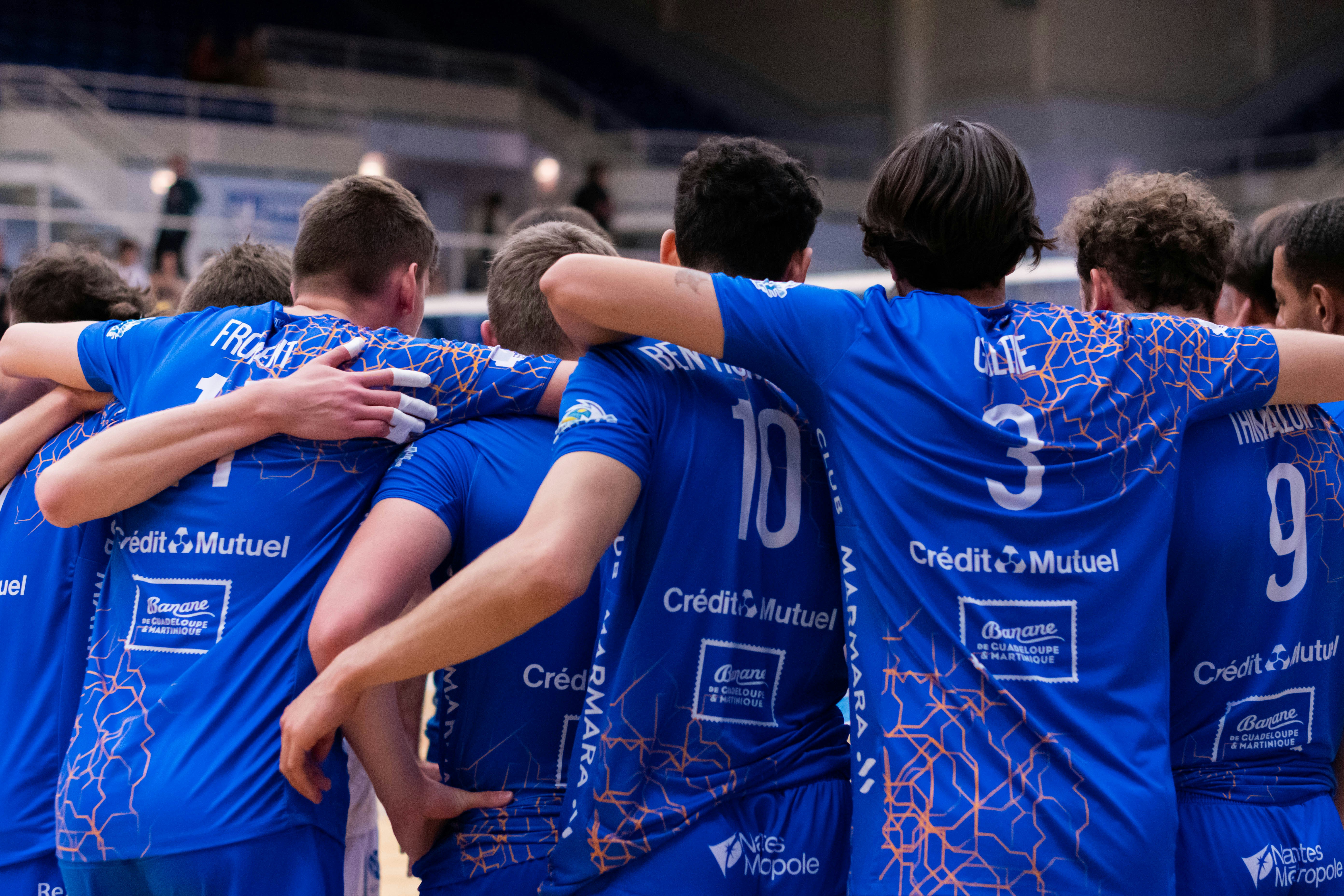 Volleyball team in blue jerseys huddling with arms interlocked during a game in an indoor arena.