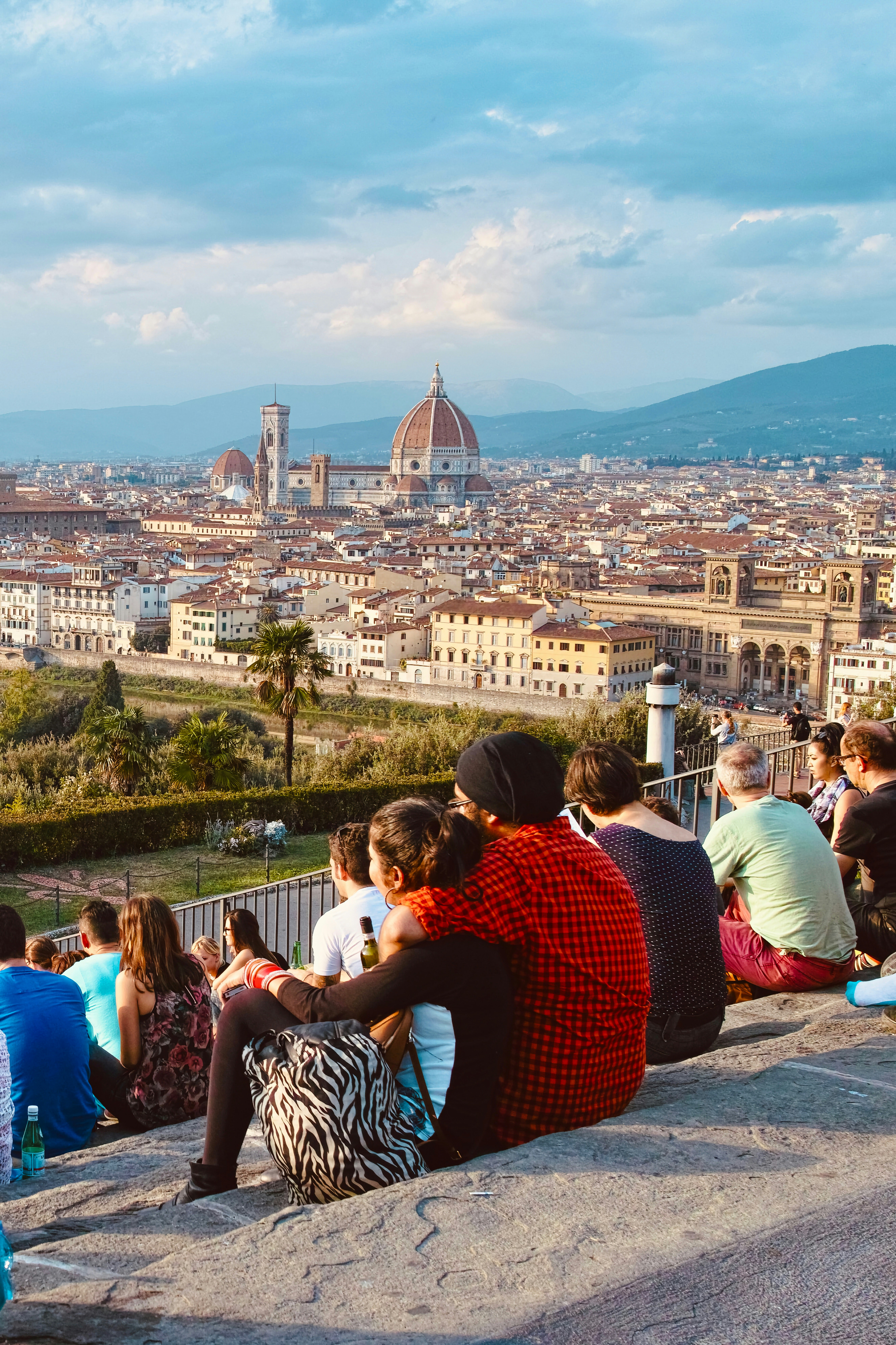 Group of people seated on a hillside, gazing at the Florence skyline with the iconic dome in the background. The warm evening light enhances the scene.