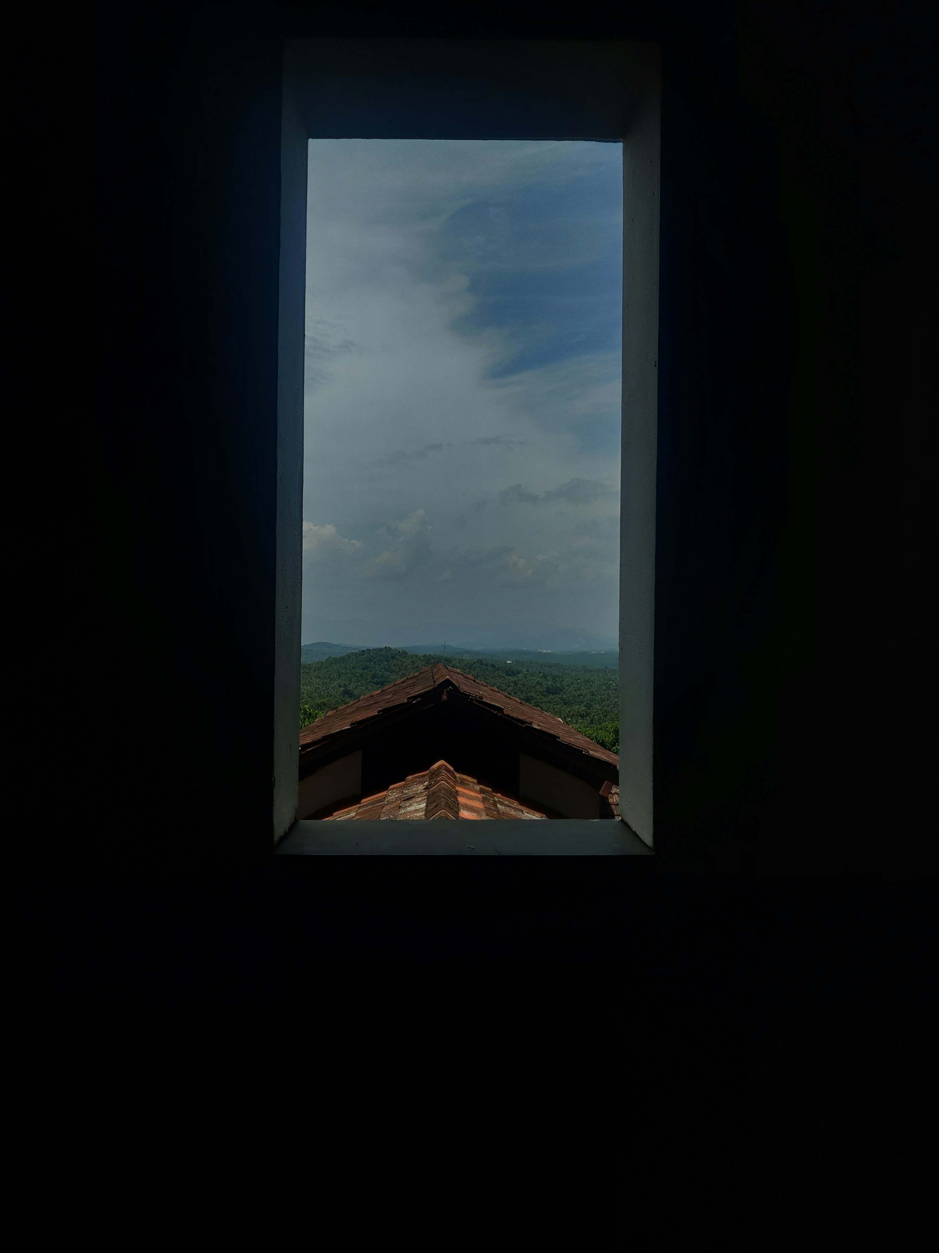 A narrow window frames a terracotta roof and a verdant horizon beneath a blue sky.