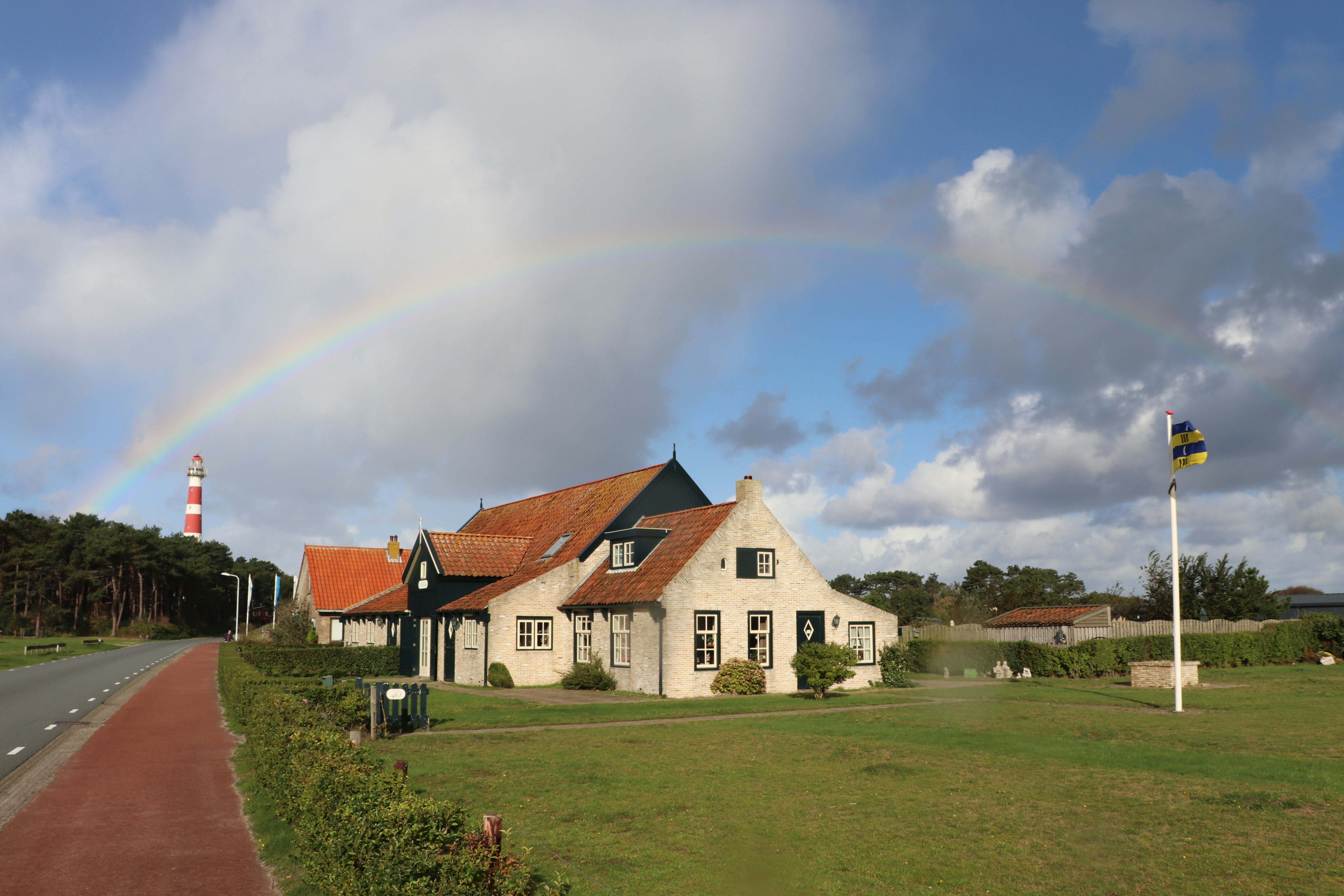 Rainbow over a white-walled cottage by a rural road with a distant lighthouse