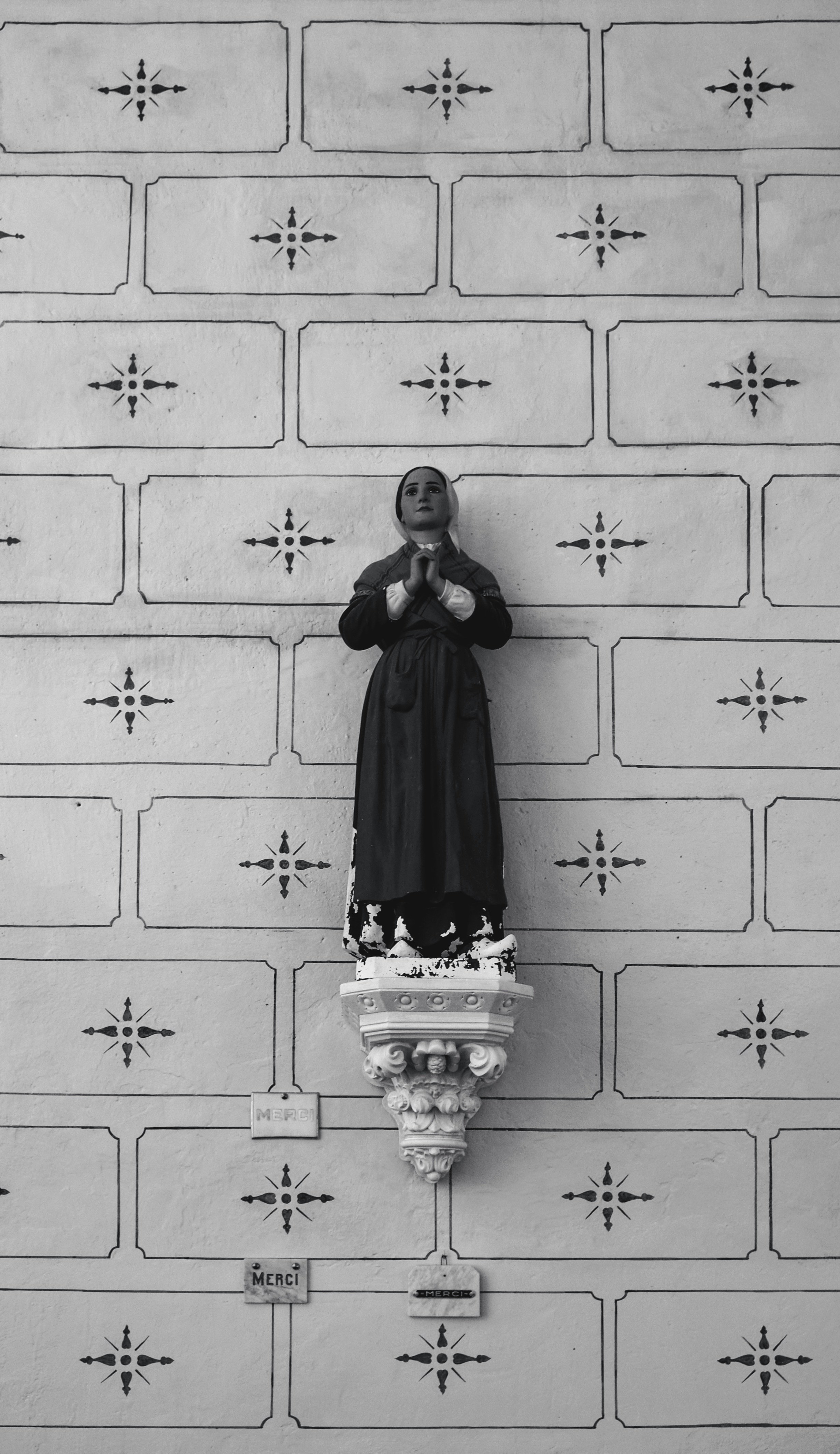 Centered Benediction: A Nun Statue Rests on a Carved Pedestal Beneath a Wall of Repeating Emblems