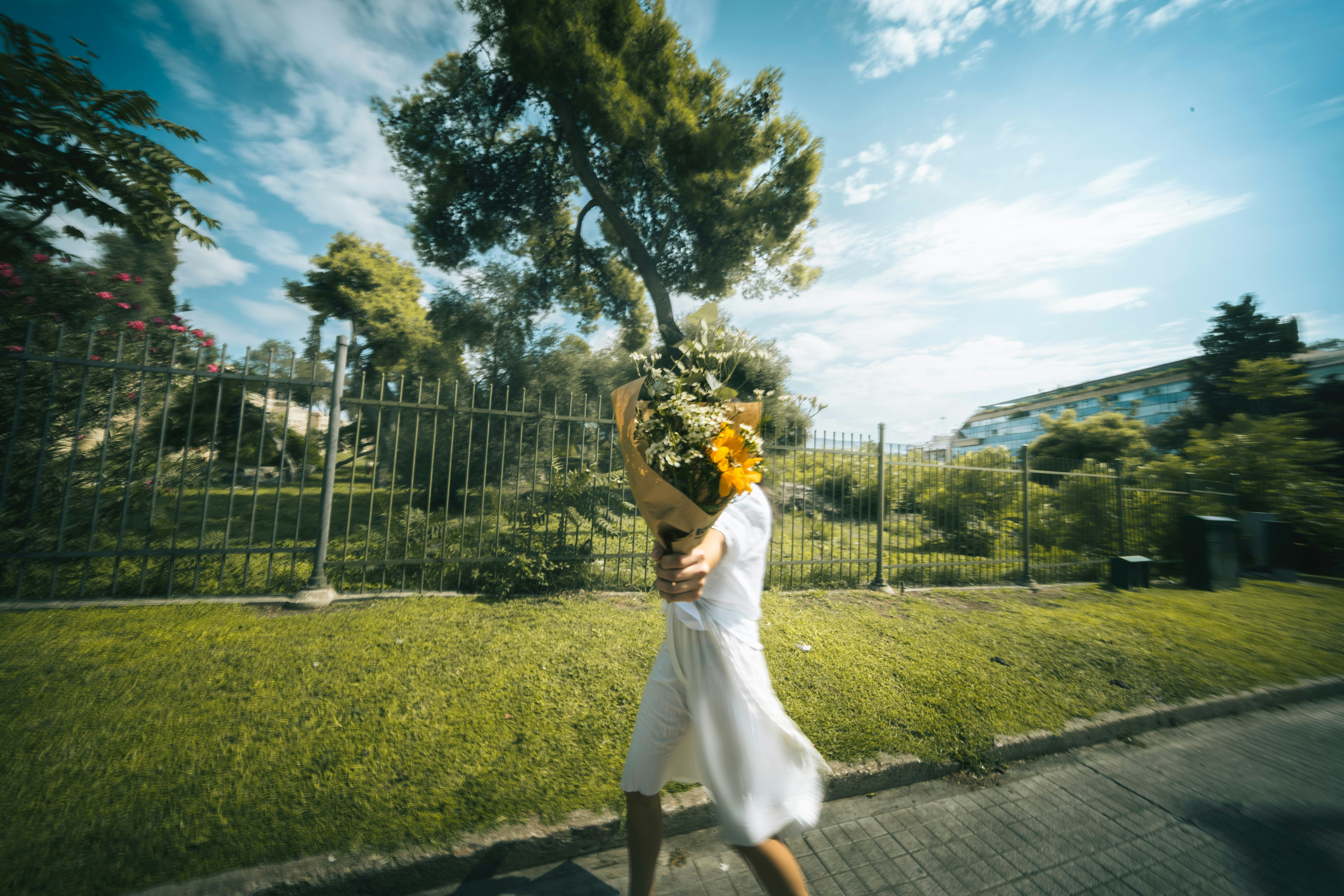 Person in white dress holding sunflowers walks energetically in a green park under a vibrant blue sky.