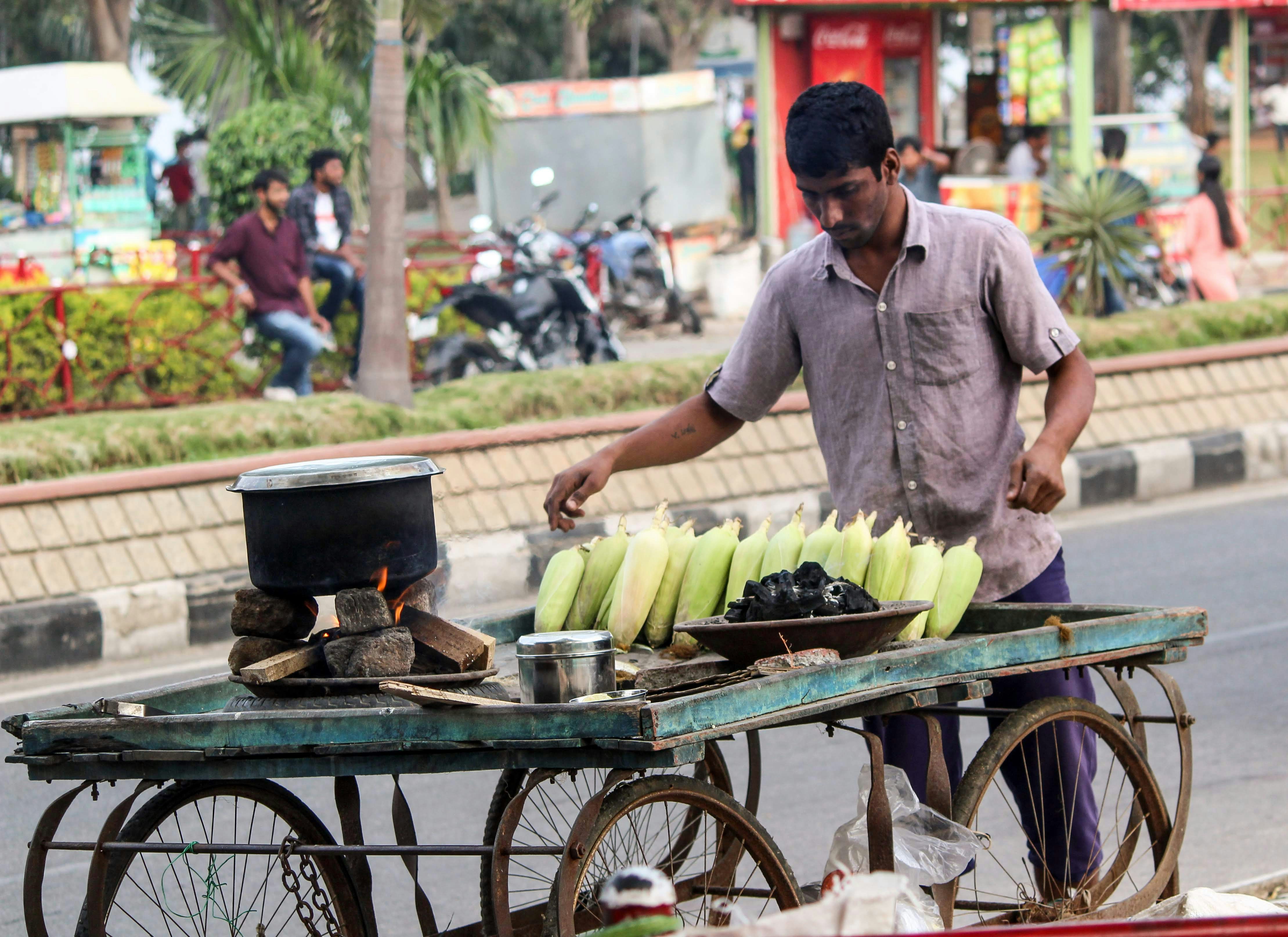 Vendor preparing roasted corn on a bustling street, surrounded by vibrant market activity.