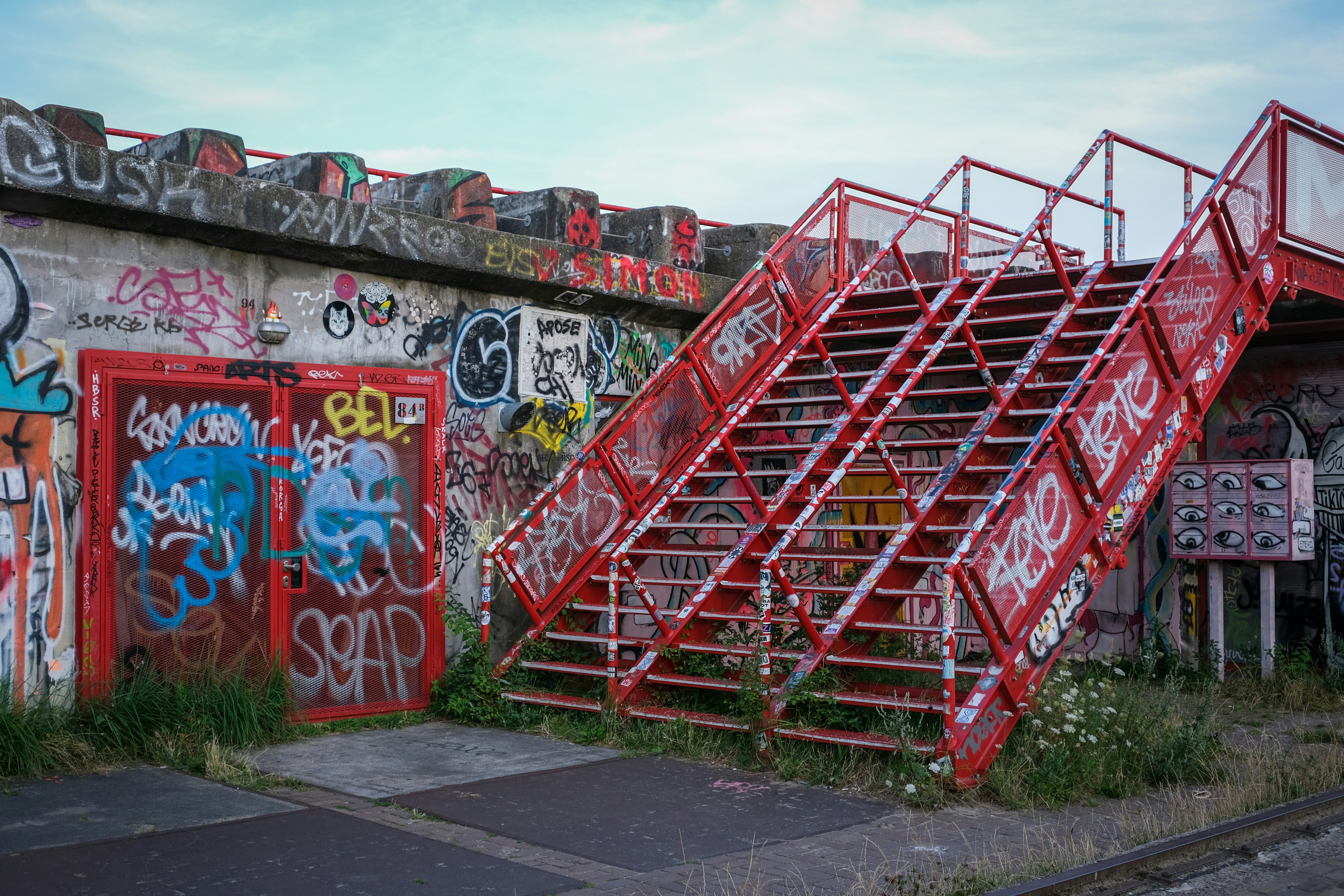Red Staircase Cuts Through a Graffiti-Stamped Concrete Corridor
