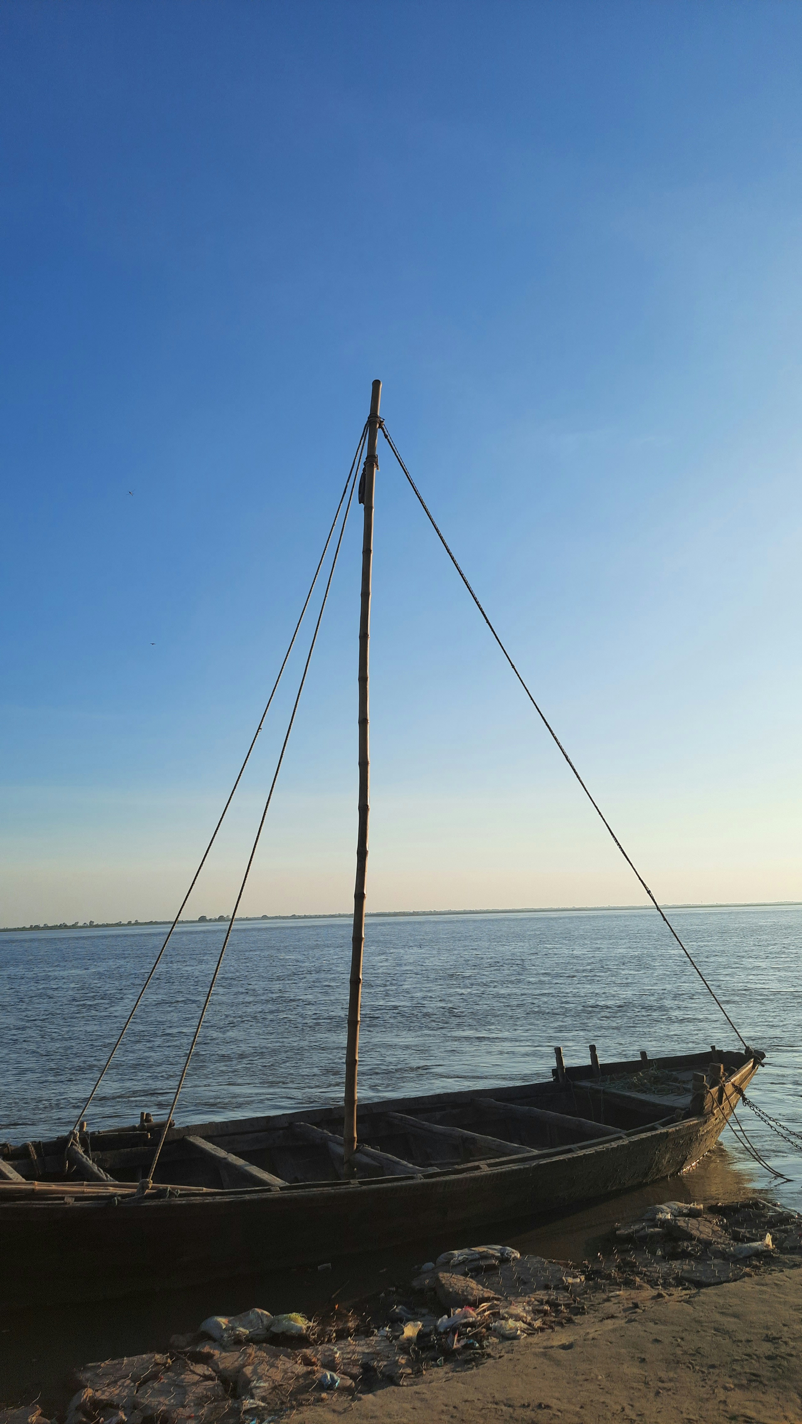 Weathered wooden boat beached along a calm shoreline with a tall central mast and taut rigging under a clear blue sky.
