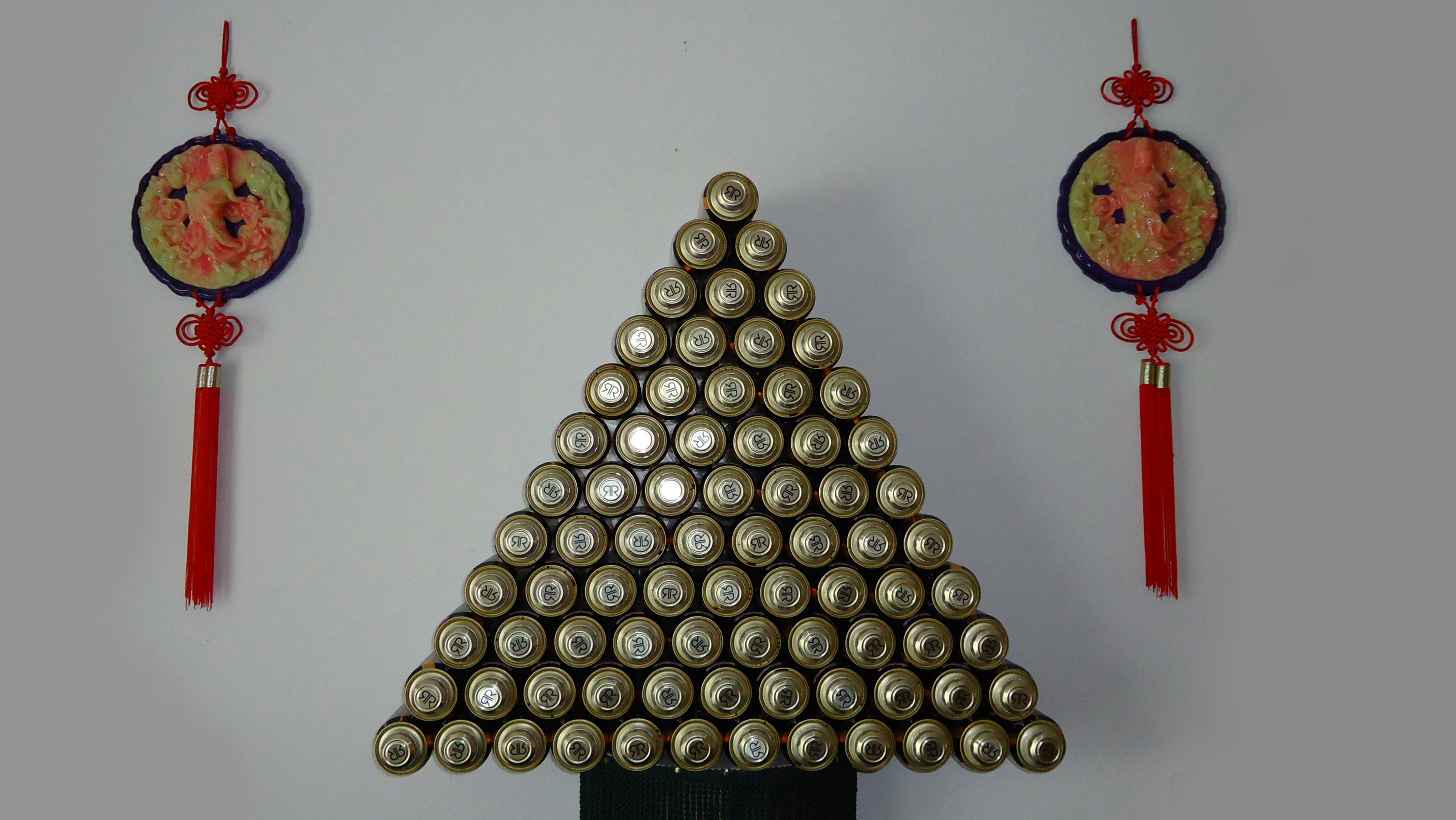 Row of metallic bottle caps arranged into a triangular pyramid on a plain white wall, flanked by circular wall ornaments with red tassels. The composition emphasizes symmetry and texture.