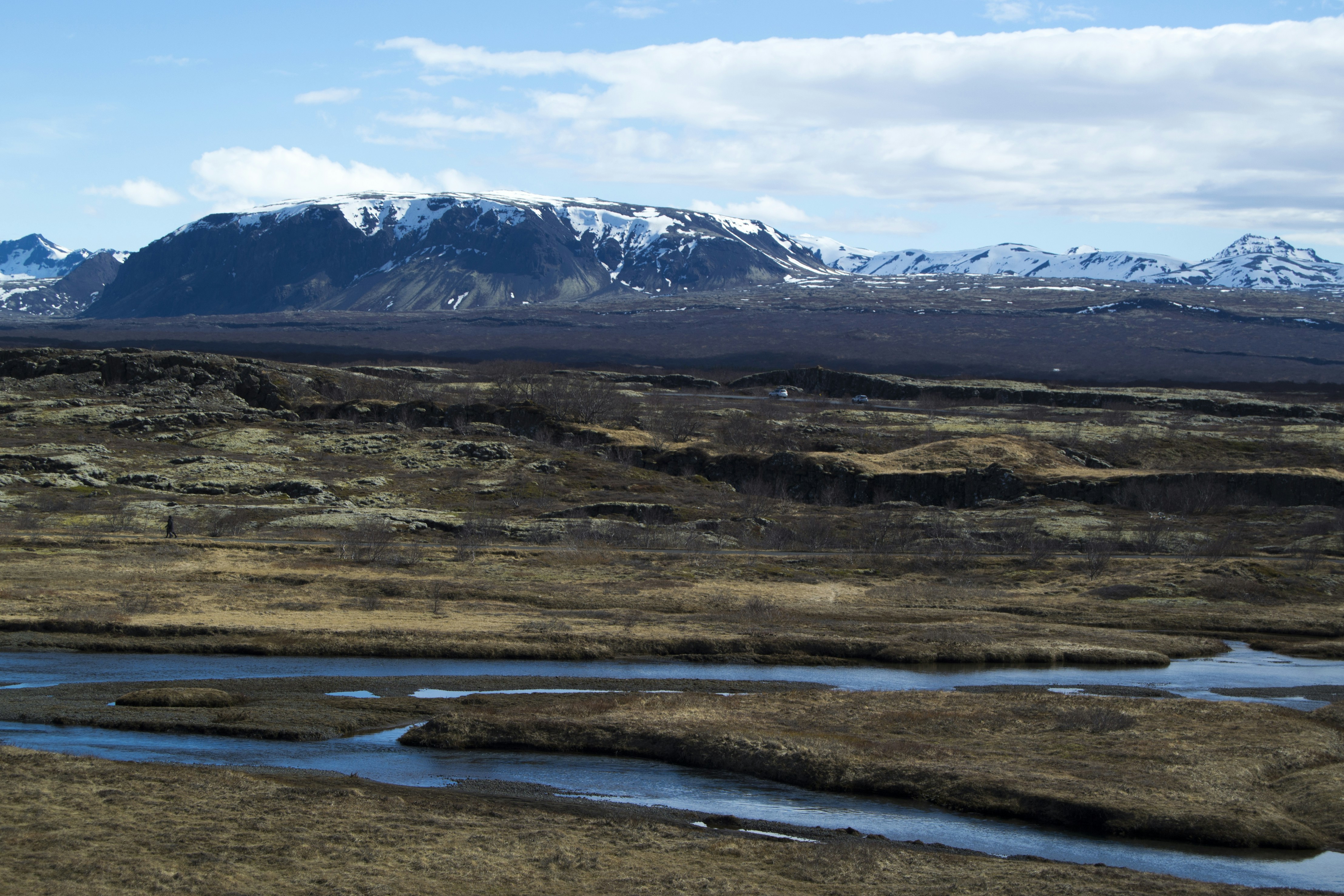 Vast landscape with winding river under snow-capped mountains and a partly cloudy sky.