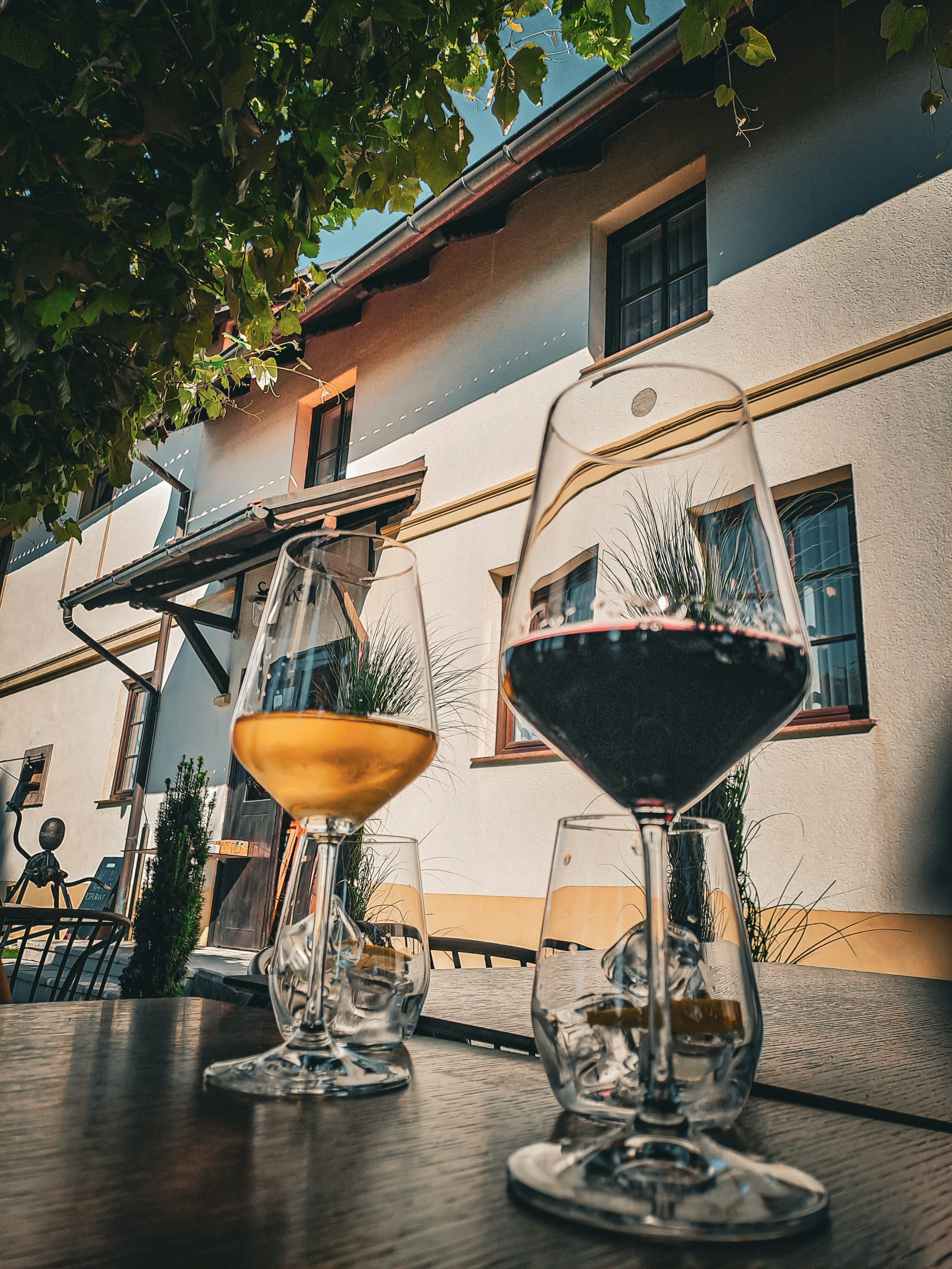 Two wine glasses sit on a wooden table in an outdoor courtyard, with a sunlit building and greenery in the background.