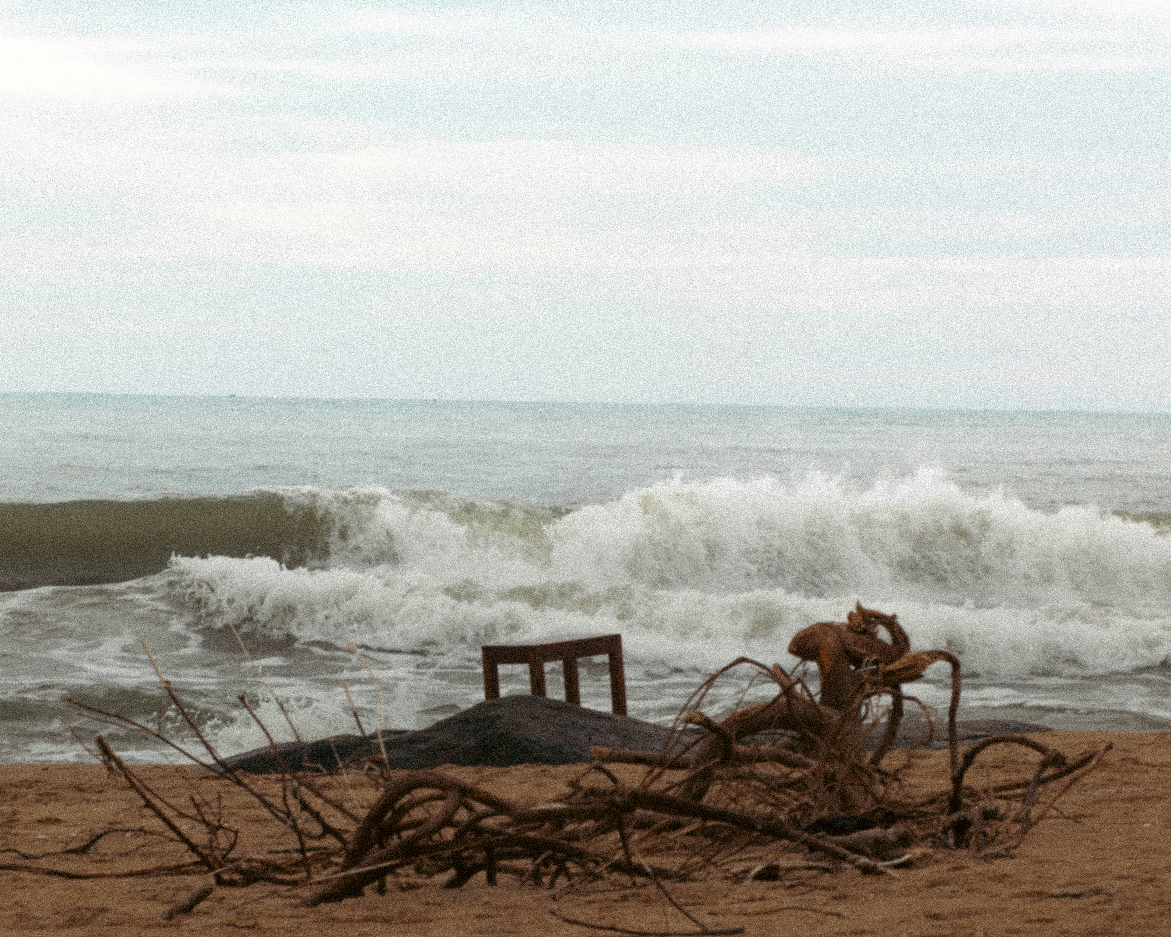A weathered chair rests on a sandy beach, surrounded by driftwood and the encroaching waves of the ocean. The scene evokes a sense of isolation and contemplation.