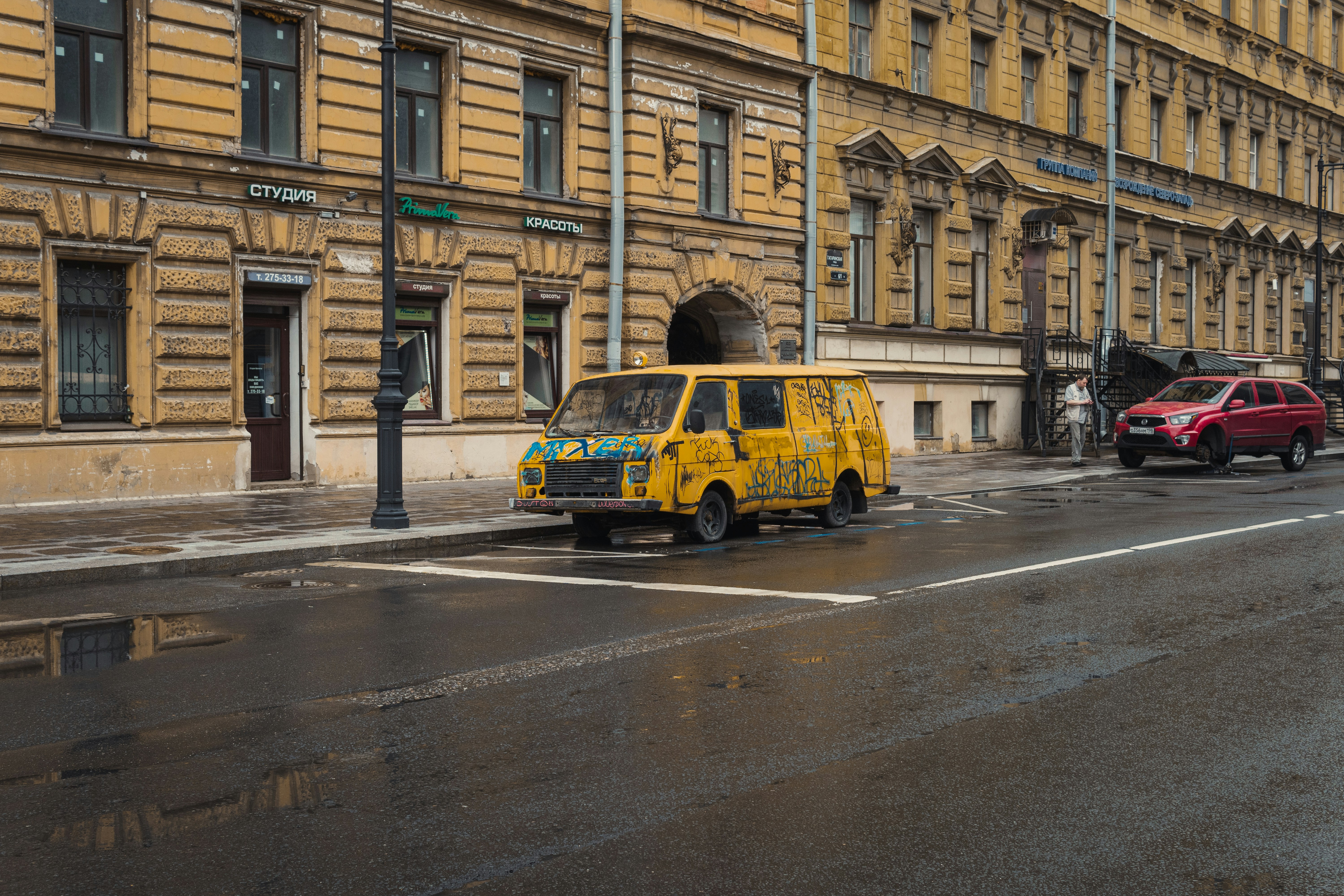 Worn yellow van halts beside a grand brick facade on a rain-soaked street