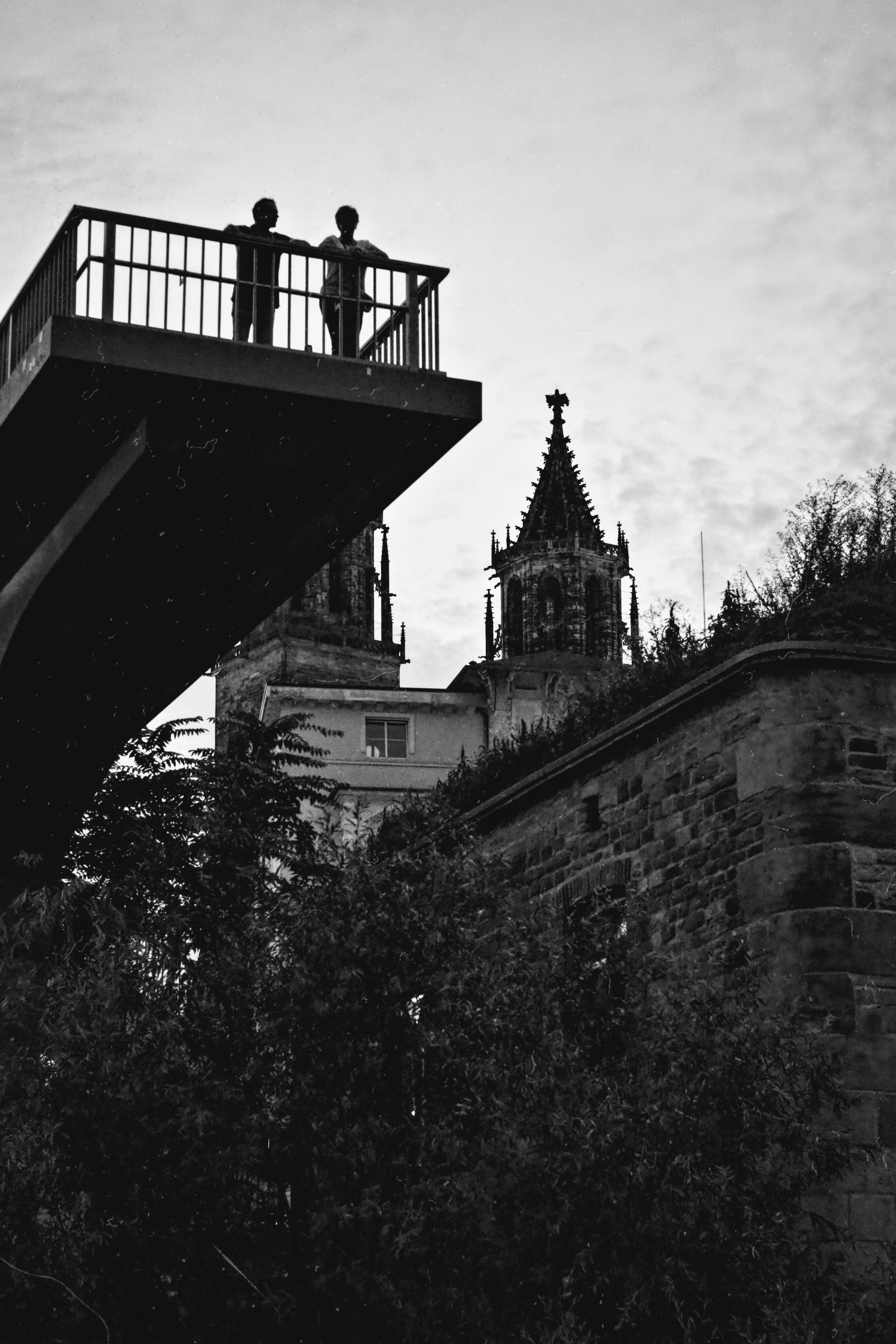Two figures stand on an elevated platform silhouetted against historic church spires.