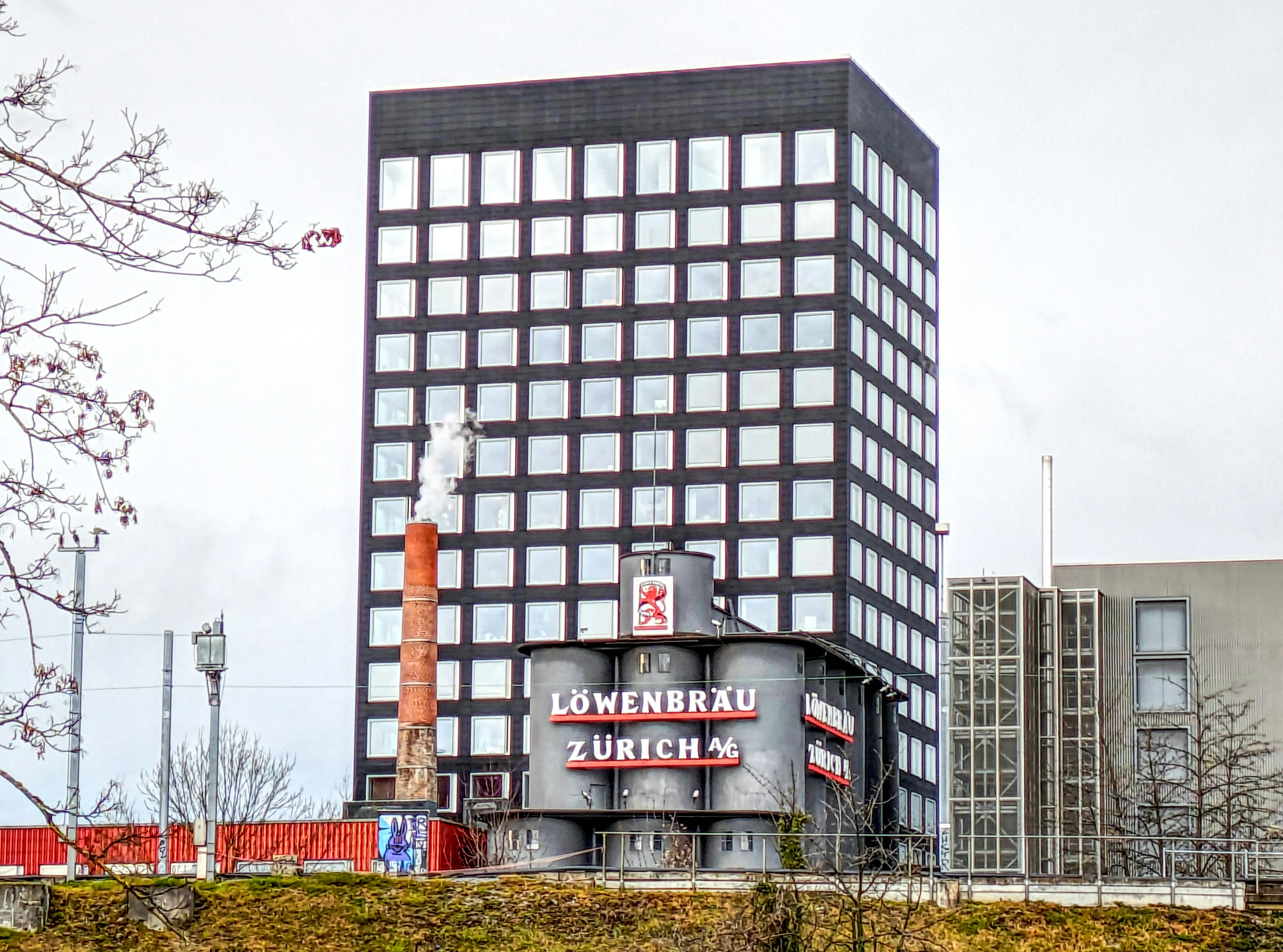 Glass-clad office tower with a grid of windows rises behind an industrial complex featuring the Löwenbräu sign and a tall smokestack. The composition emphasizes the contrast between corporate architecture and industrial heritage.