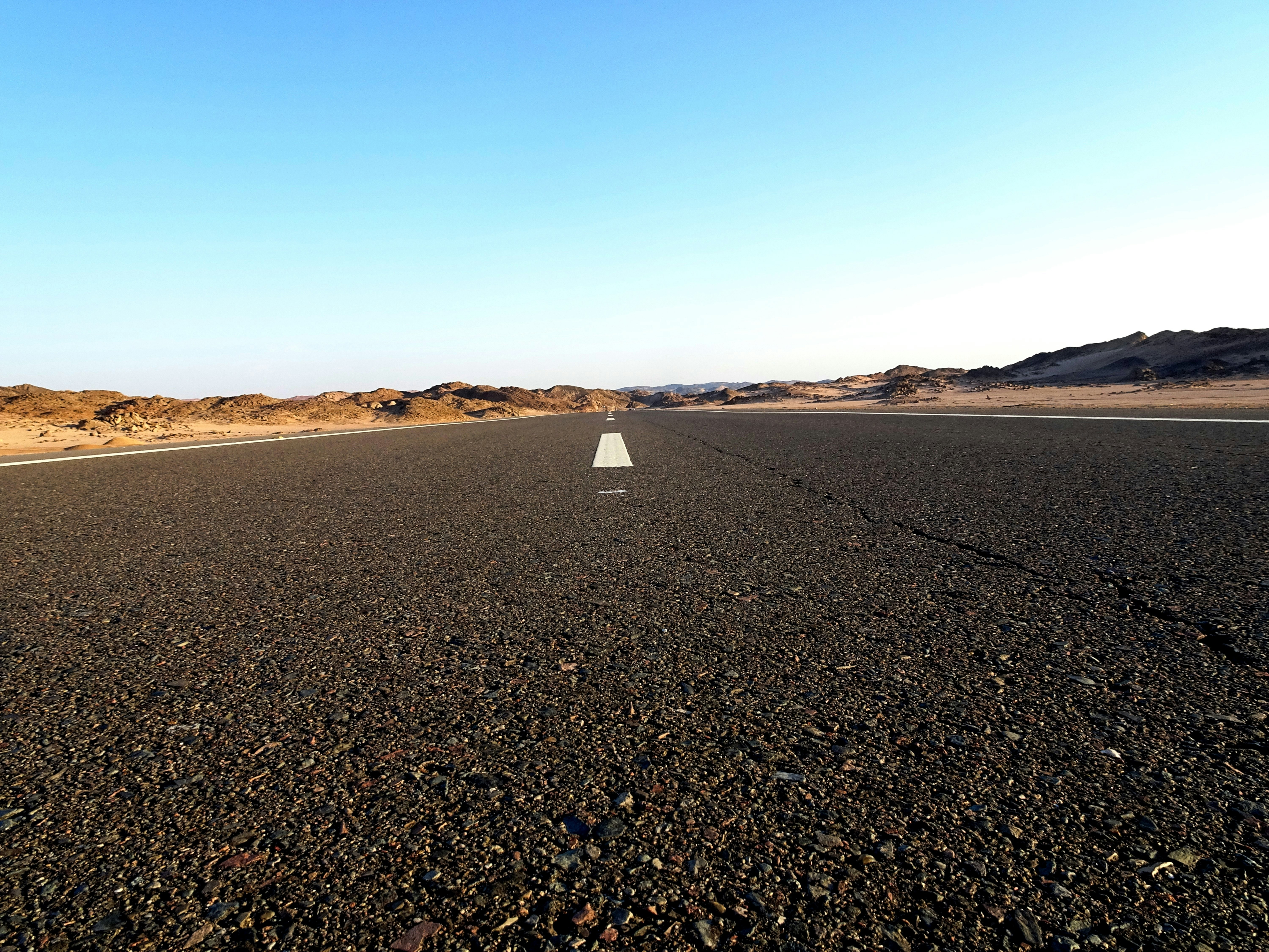 Straightaway road stretches toward the horizon with a single white center line under a bright blue sky, surrounded by arid dunes and texture-rich asphalt.
