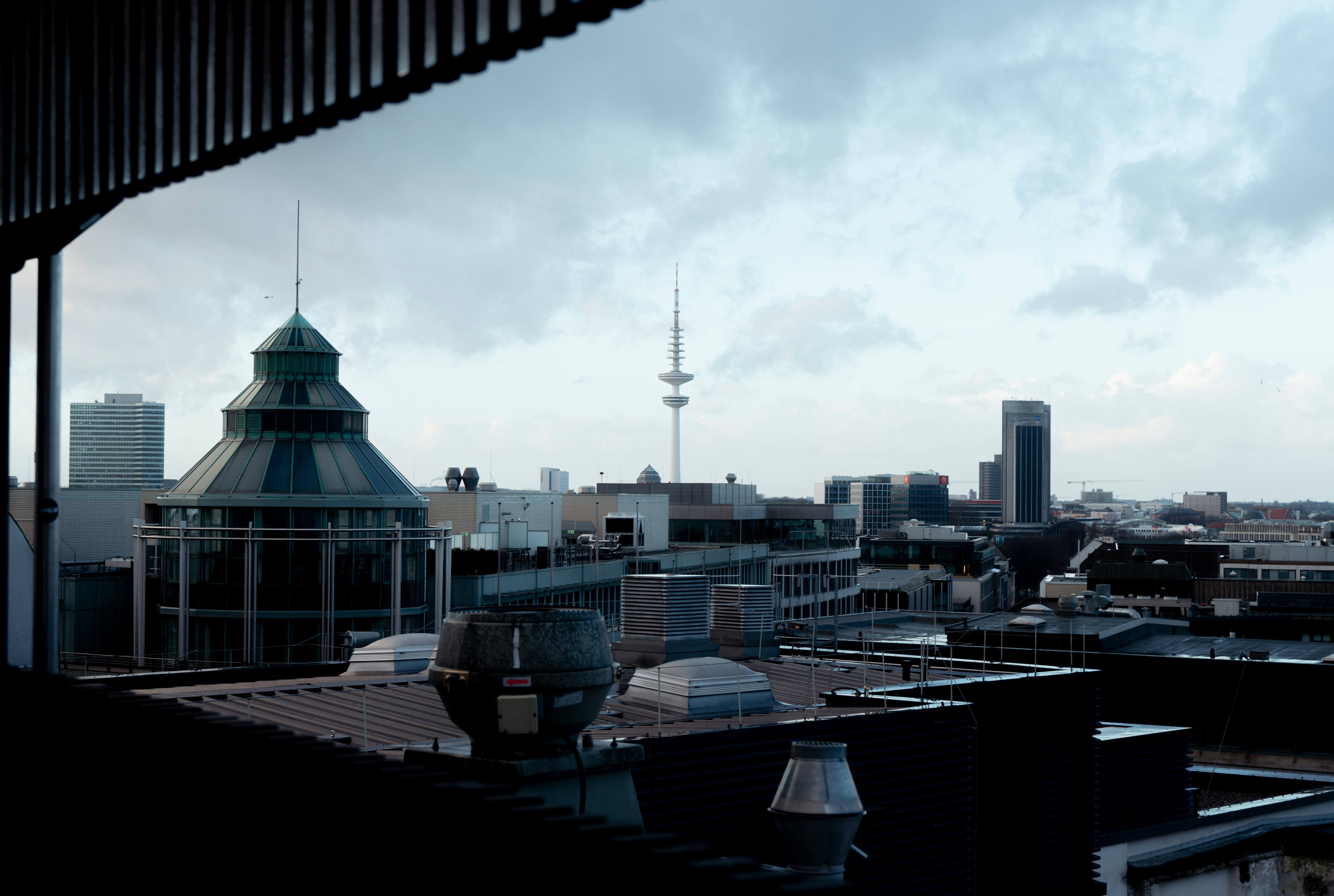 Urban skyline view captured from a shaded rooftop, featuring geometric rooftops, a distant communication tower, and a moody sky.