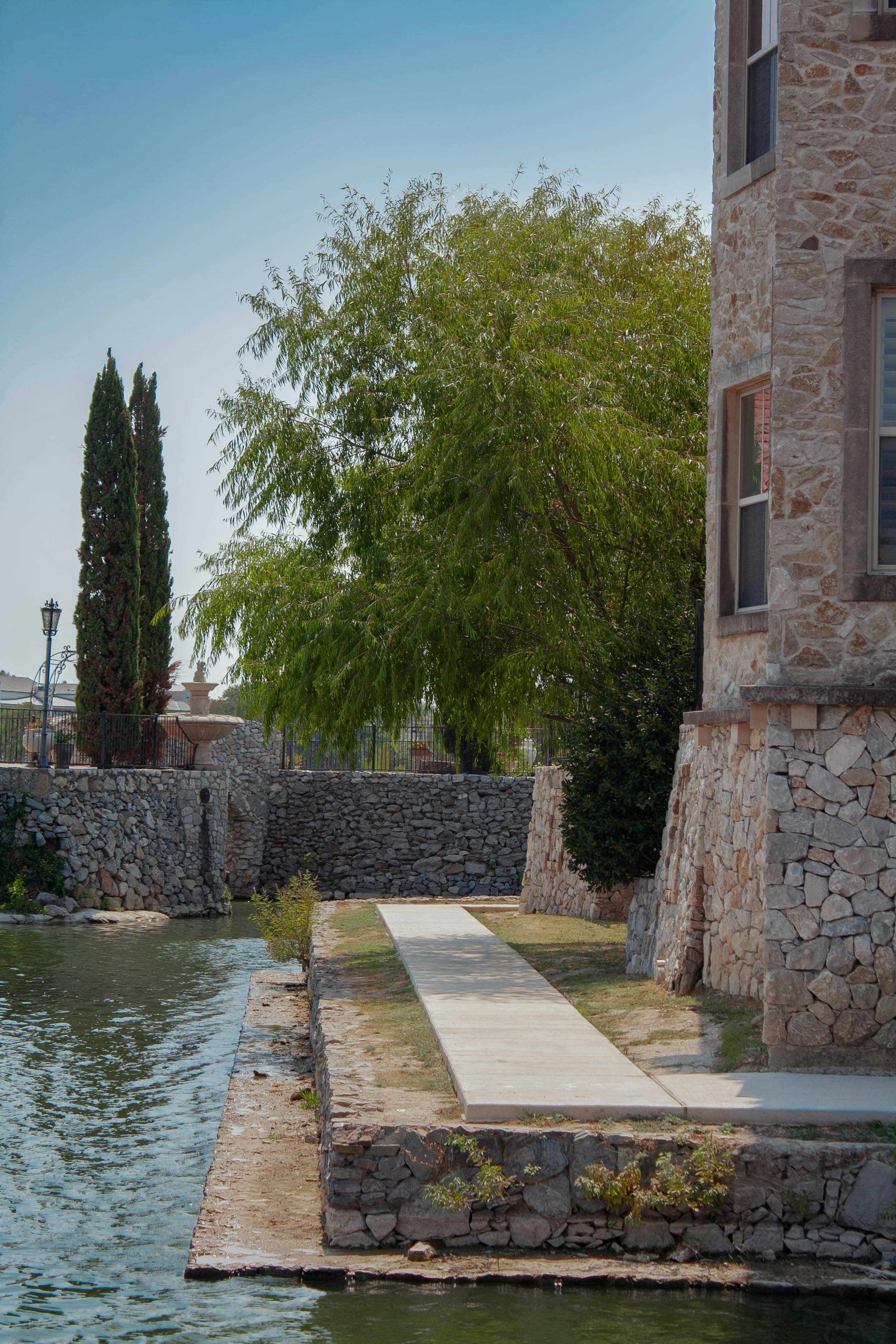 A tranquil riverside scene featuring a stone pathway alongside lush greenery and a quaint building. The calm water reflects the surrounding nature.