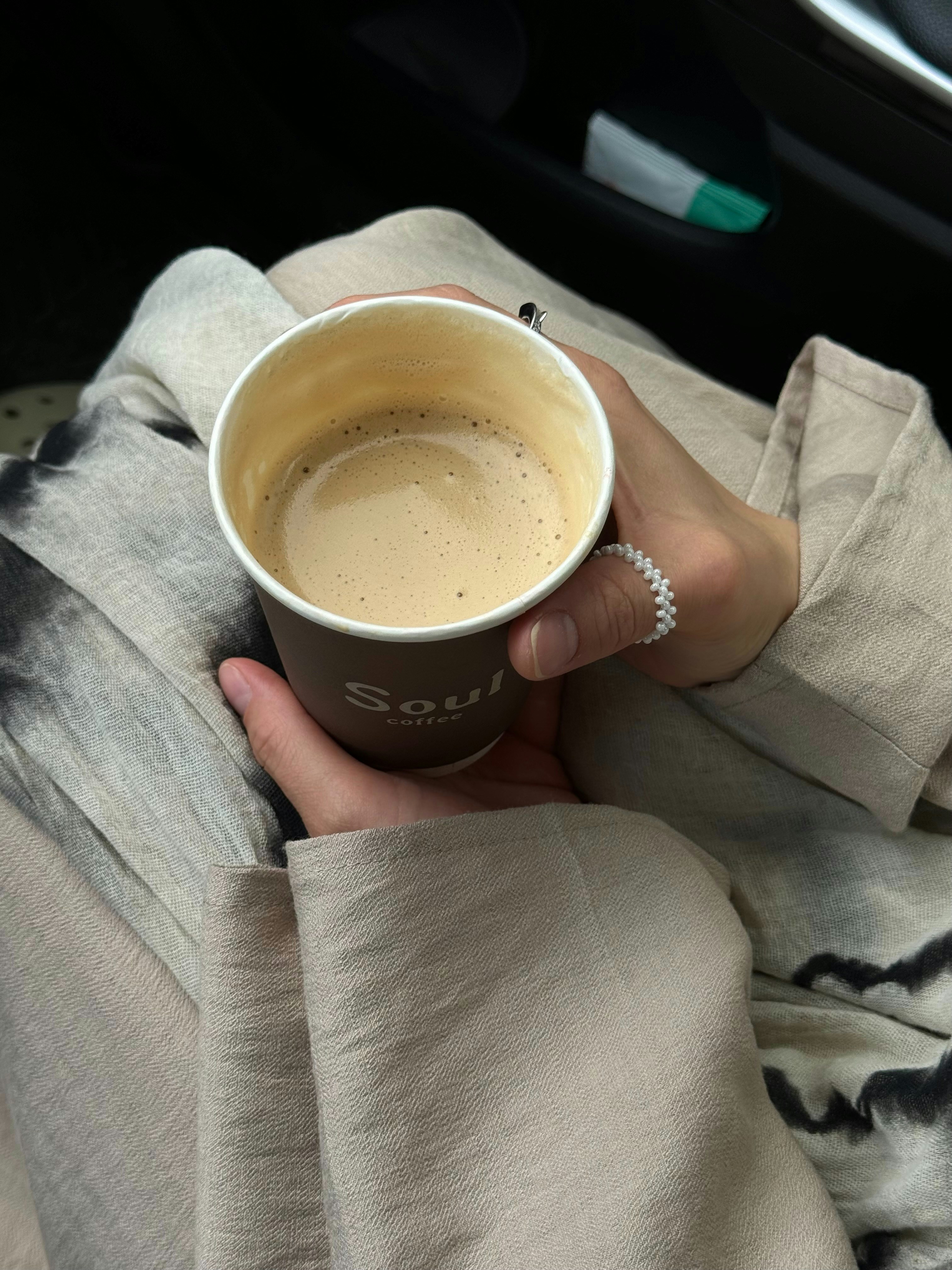 Close-up of a creamy latte cradled in hands, with a beaded ring and beige scarf, inside a car.