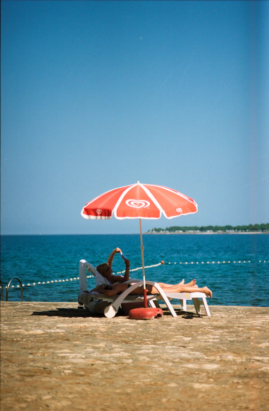 A person reclines on a beach lounger under a red umbrella beside a calm blue sea, with a distant shoreline on the horizon. Bright midday light and a wide open sky fill the frame.