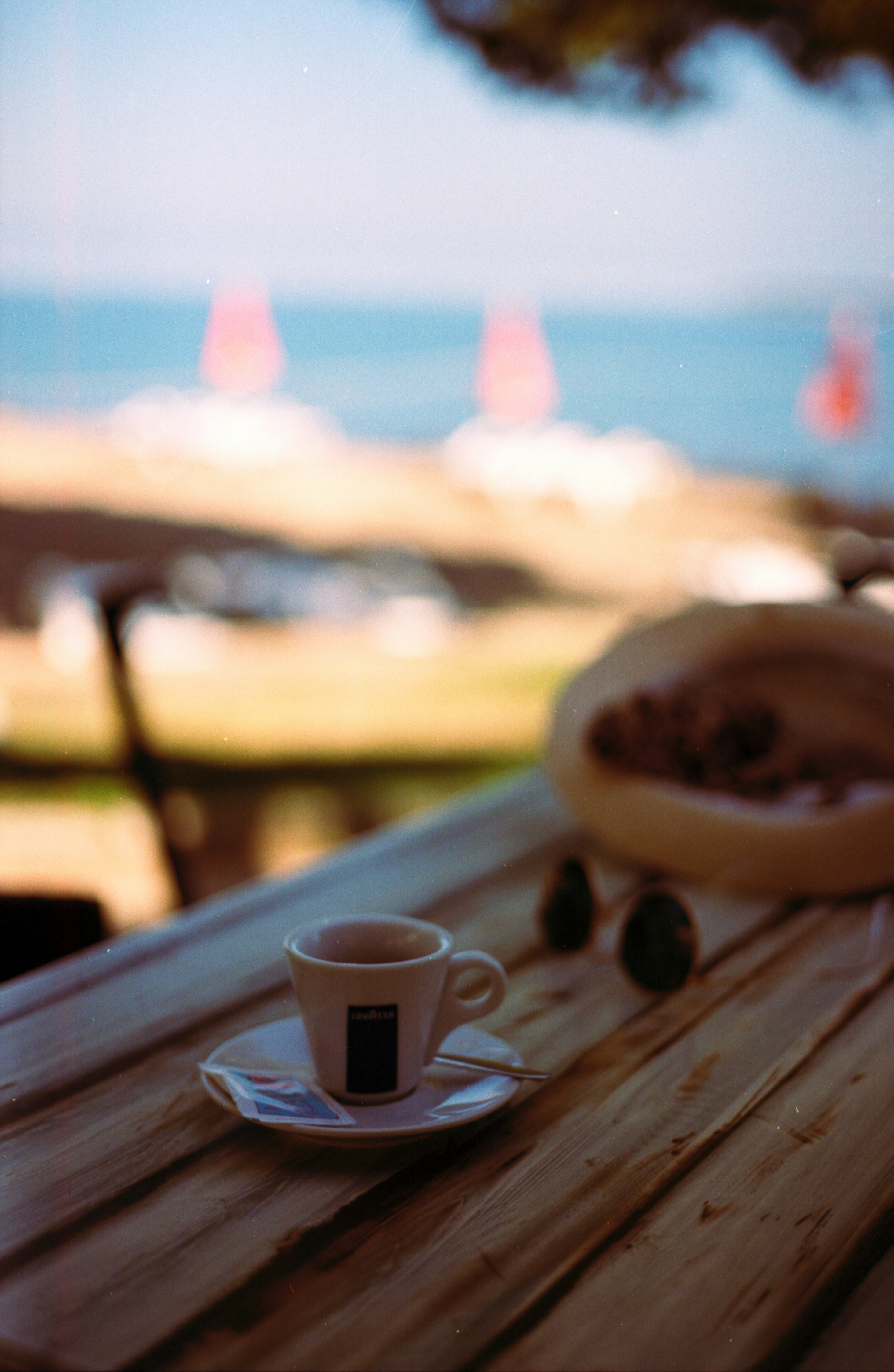 Soft-focus beach cafe scene with a tiny espresso cup on a weathered wooden table, a pastry beside it, and blurred umbrellas and sea in the background.