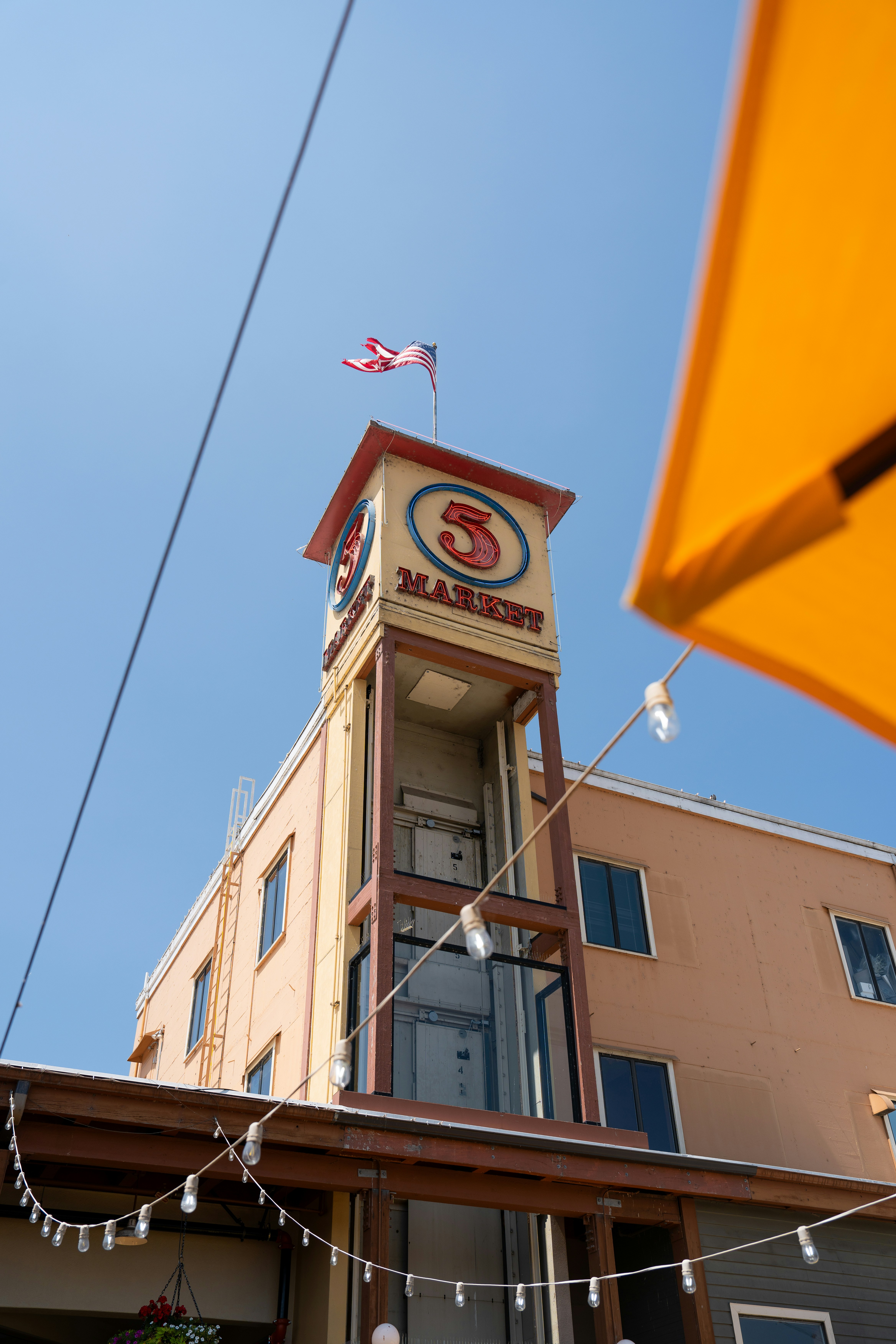 Clock Tower on a Sunny Market Day, Framed by String Lights and an Orange Canopy