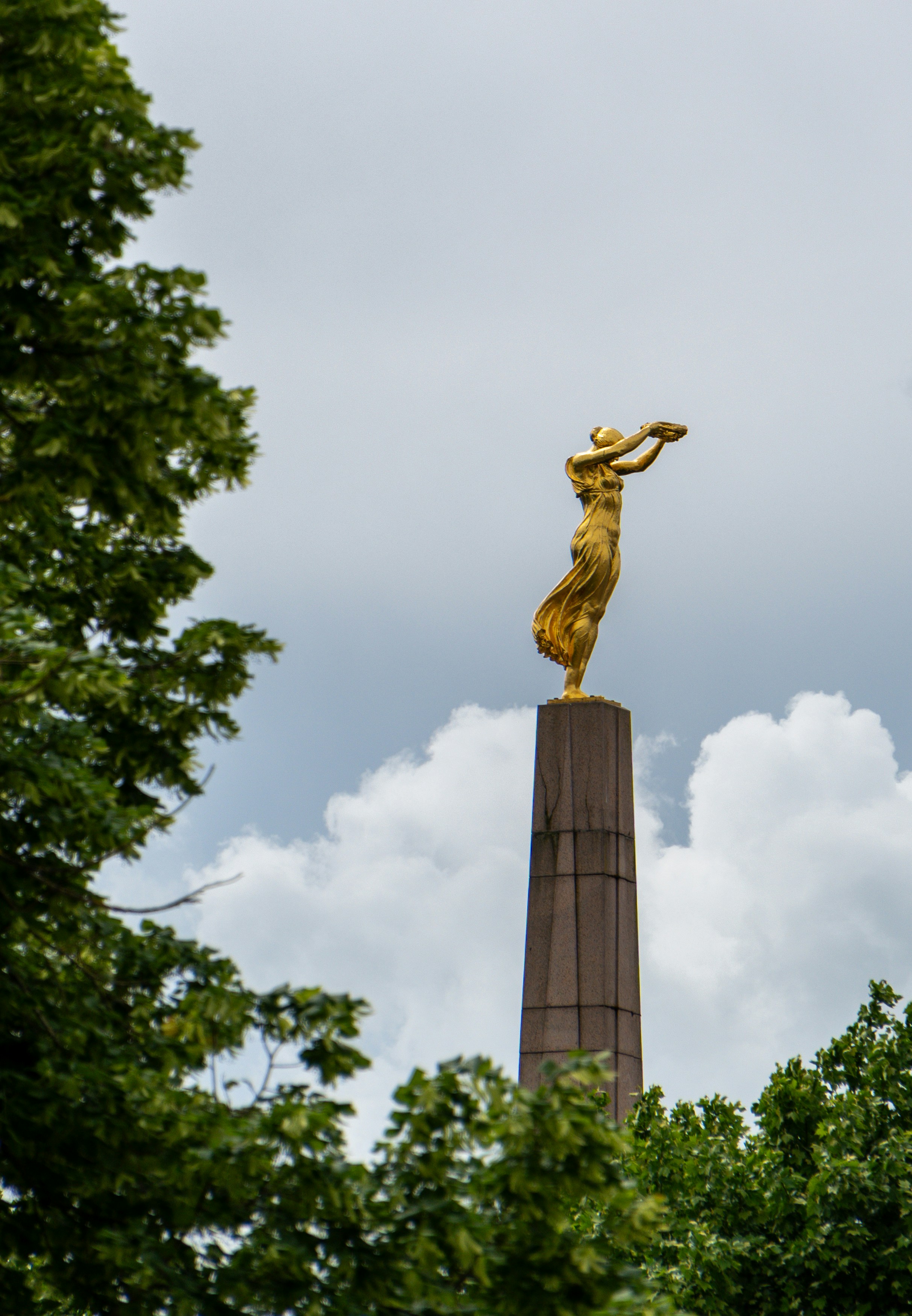 Gilded Sentinel on a Tall Obelisk Amid Sky and Trees