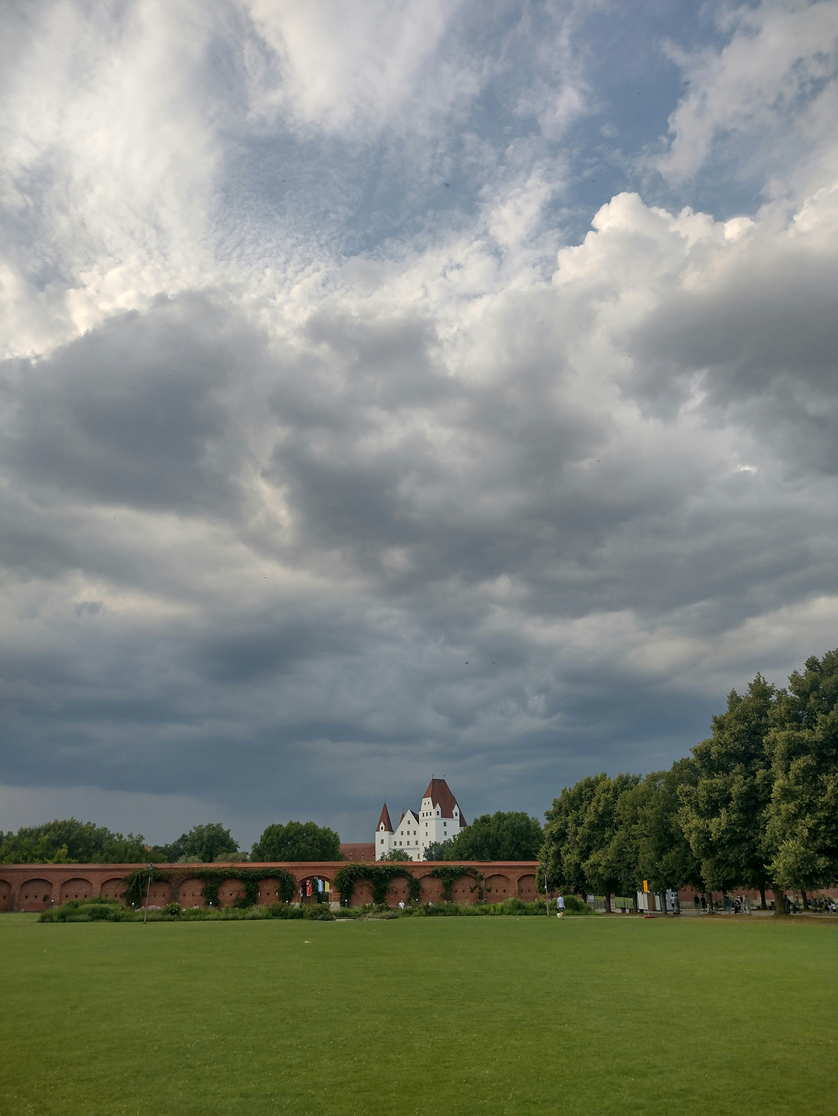 Expansive cloudy sky over a green field with distant historic architecture and trees.