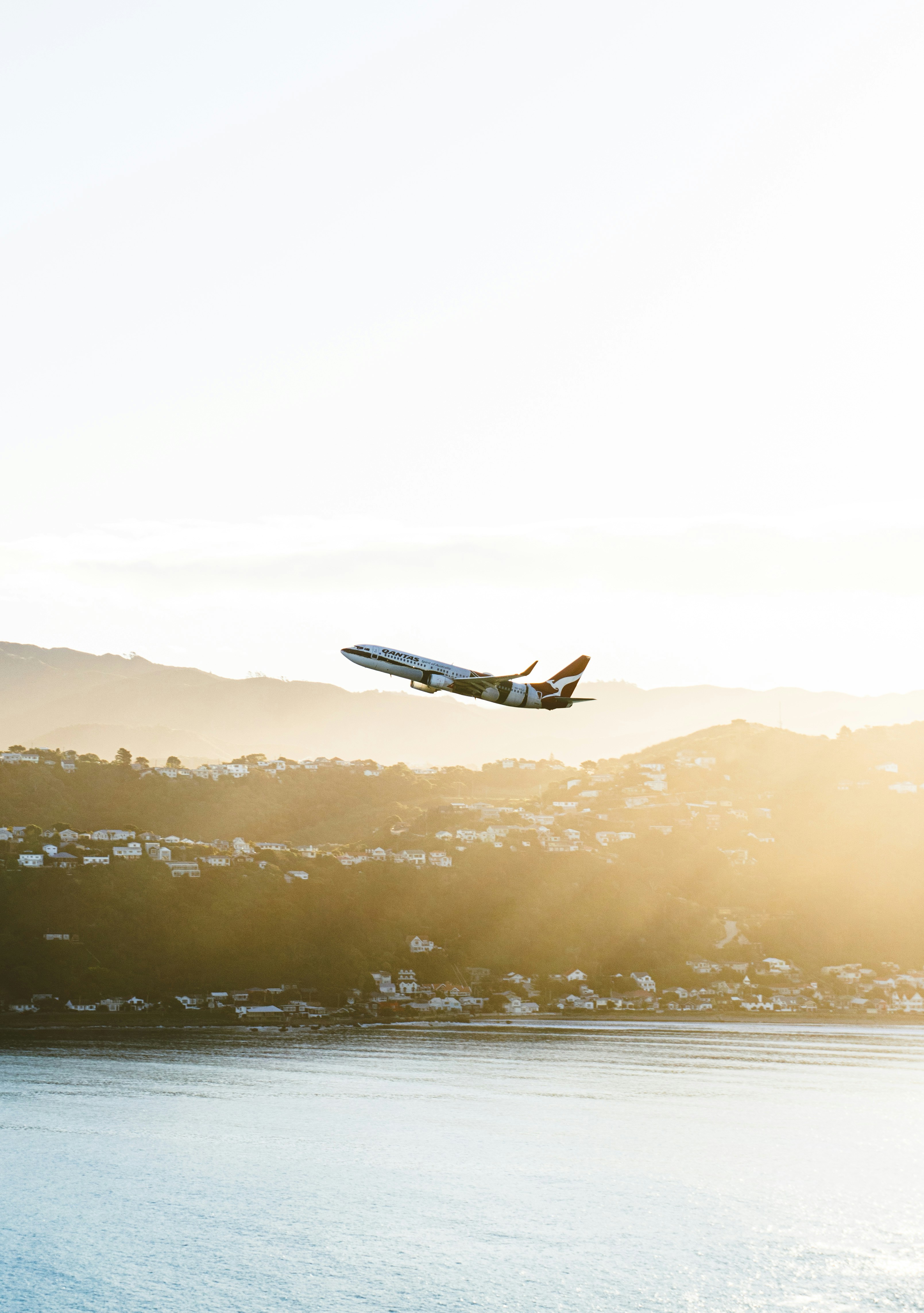 Commercial airplane ascending over a serene landscape at sunset, with gentle hills and a glimmering bay below.