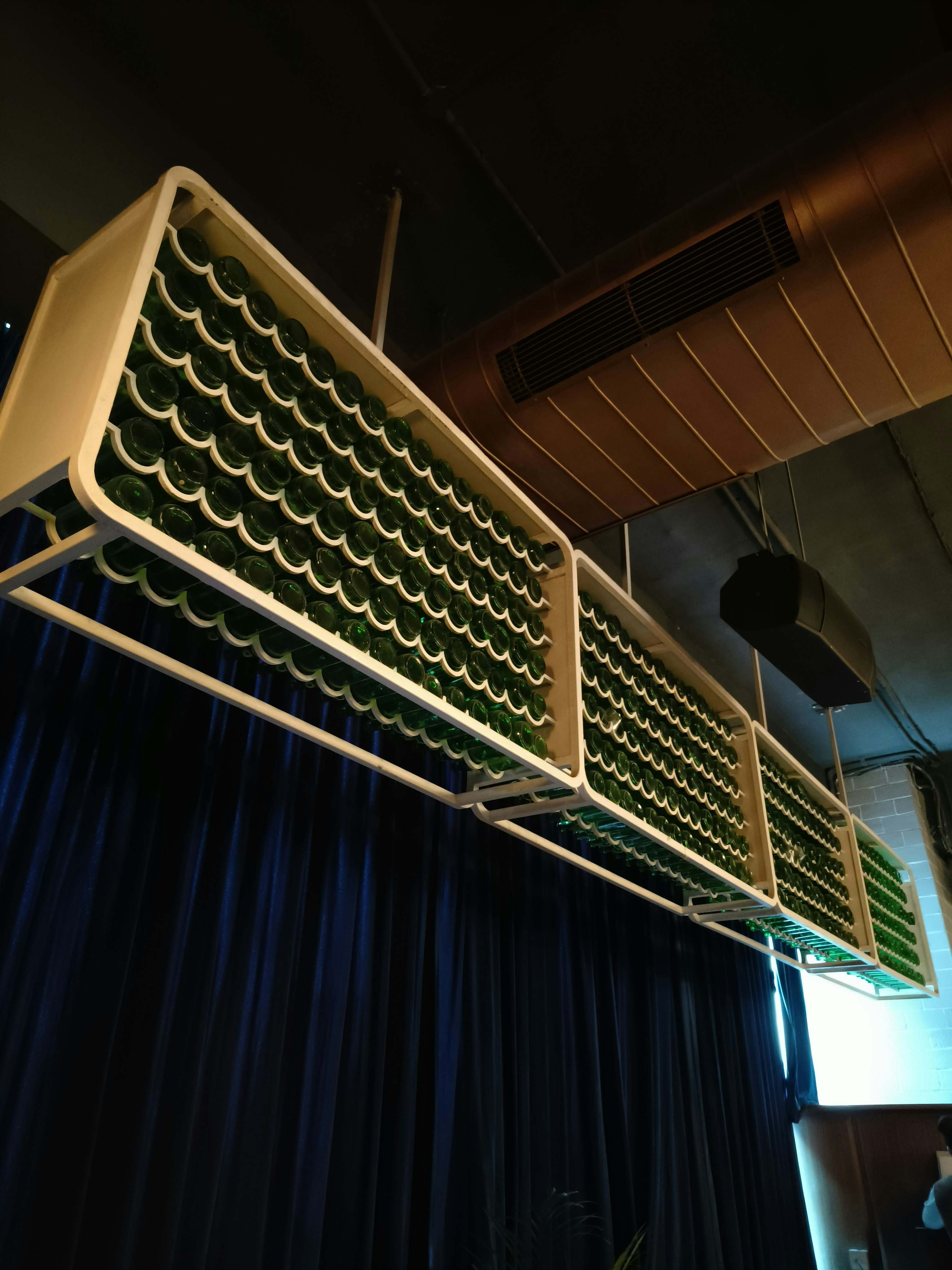 Hanging racks of green bottles above a dim stage with blue curtains, viewed from an angled ceiling perspective.