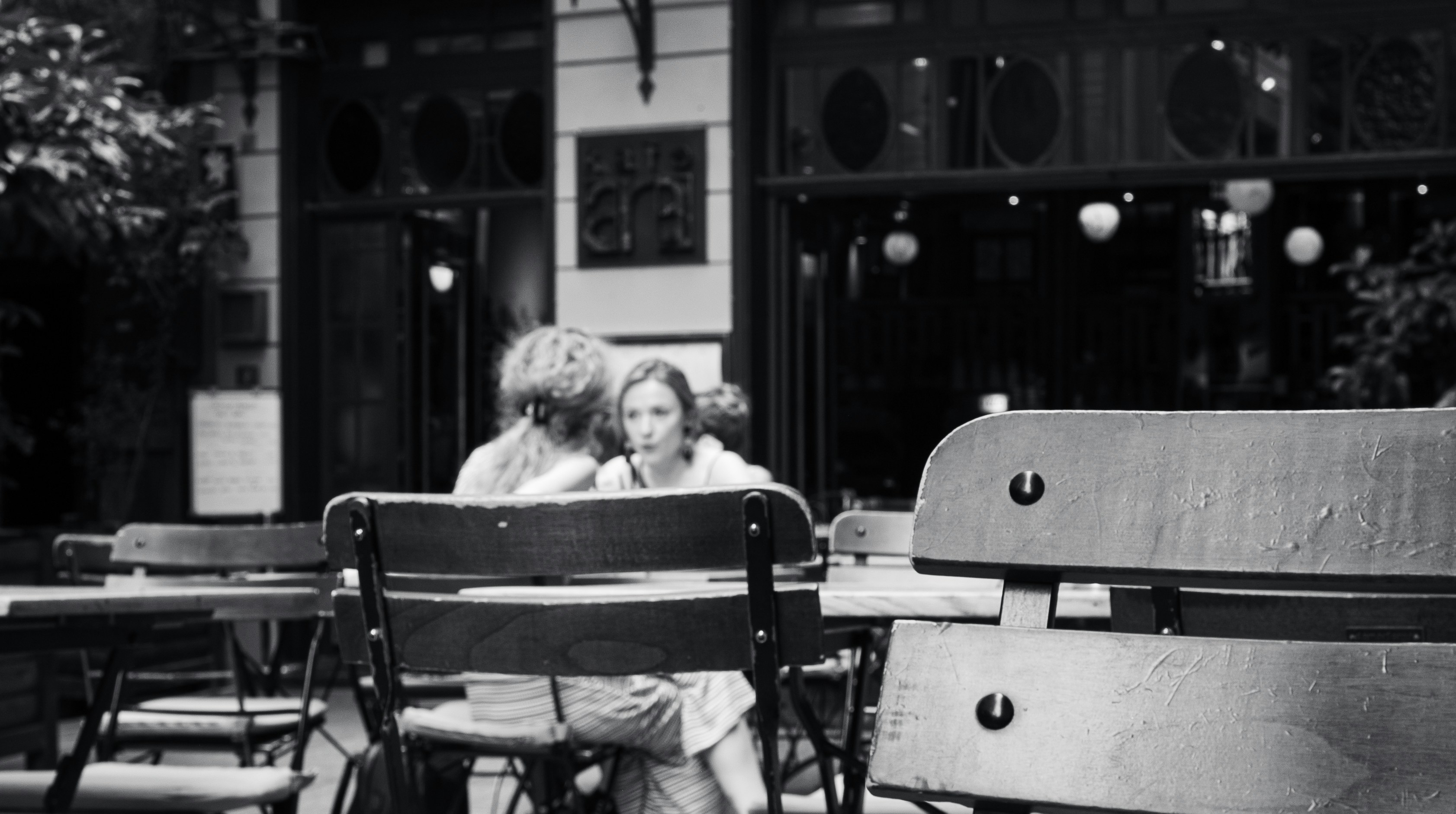 Two people converse at an outdoor café table, with blurred foreground chairs and a softly focused backdrop.